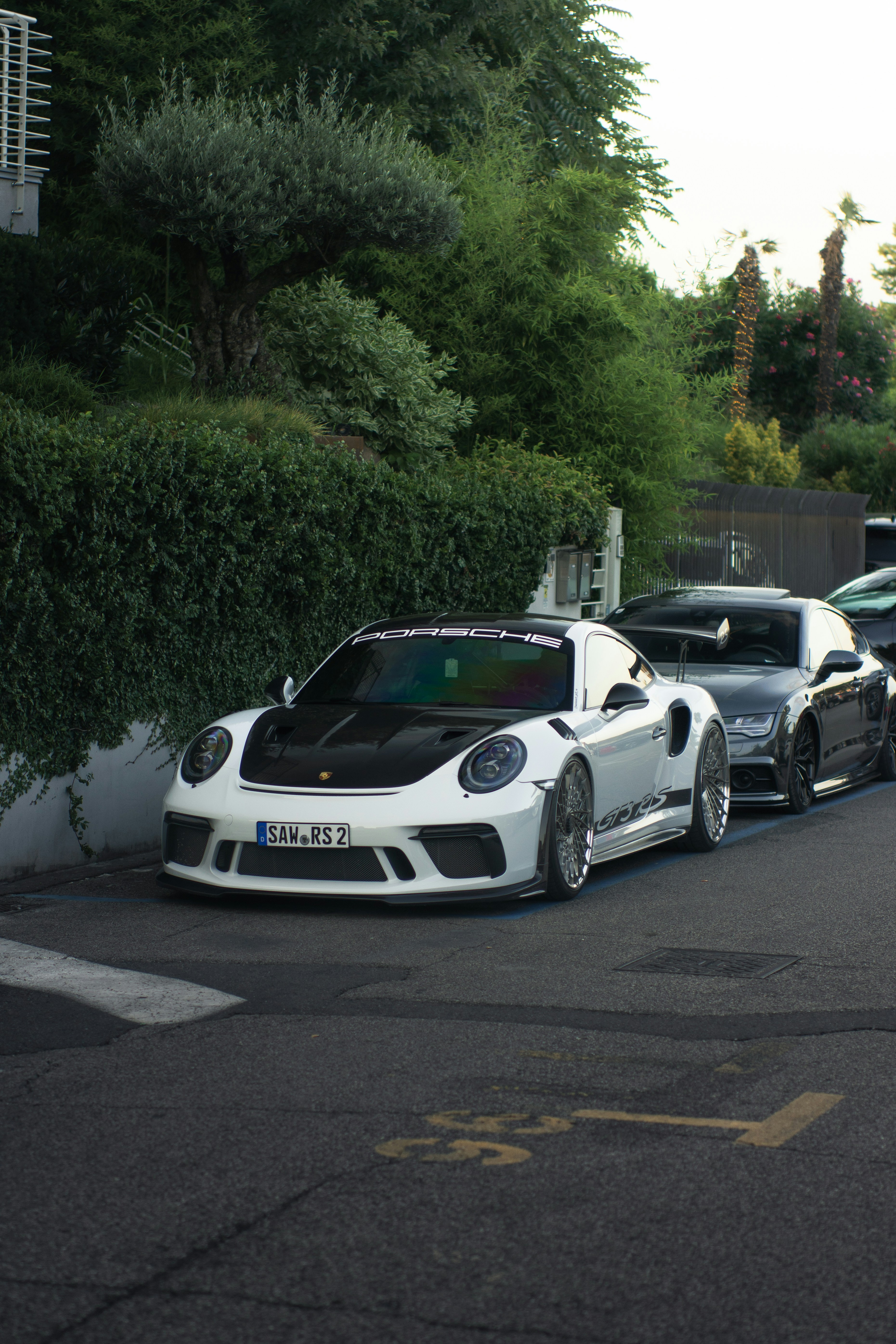 A sleek white Porsche 911 GT2 RS parked alongside a black sports car, framed by lush greenery and urban architecture.