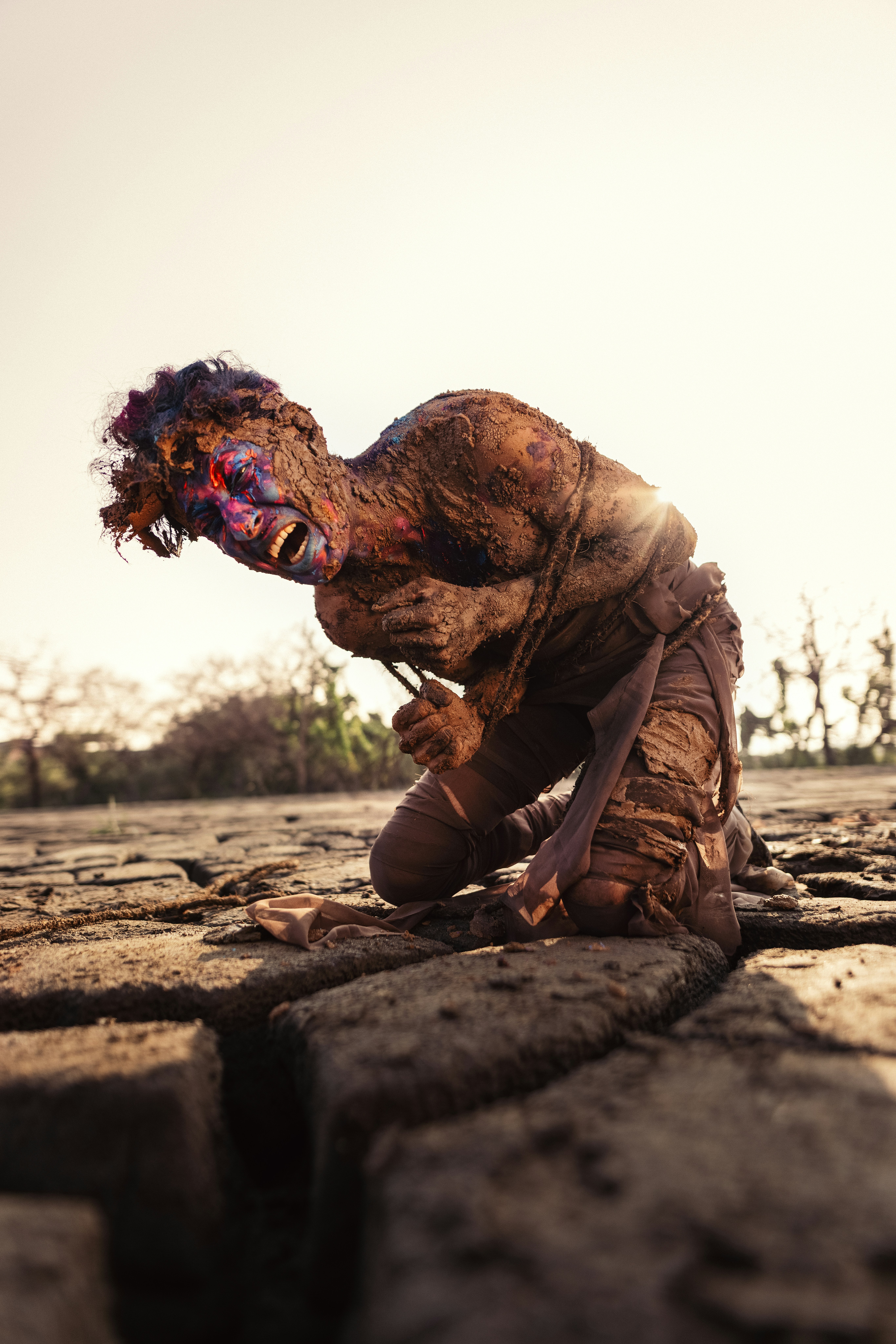 A person covered in mud, expressing intense emotion, kneels on cracked earth, symbolizing struggle and resilience.