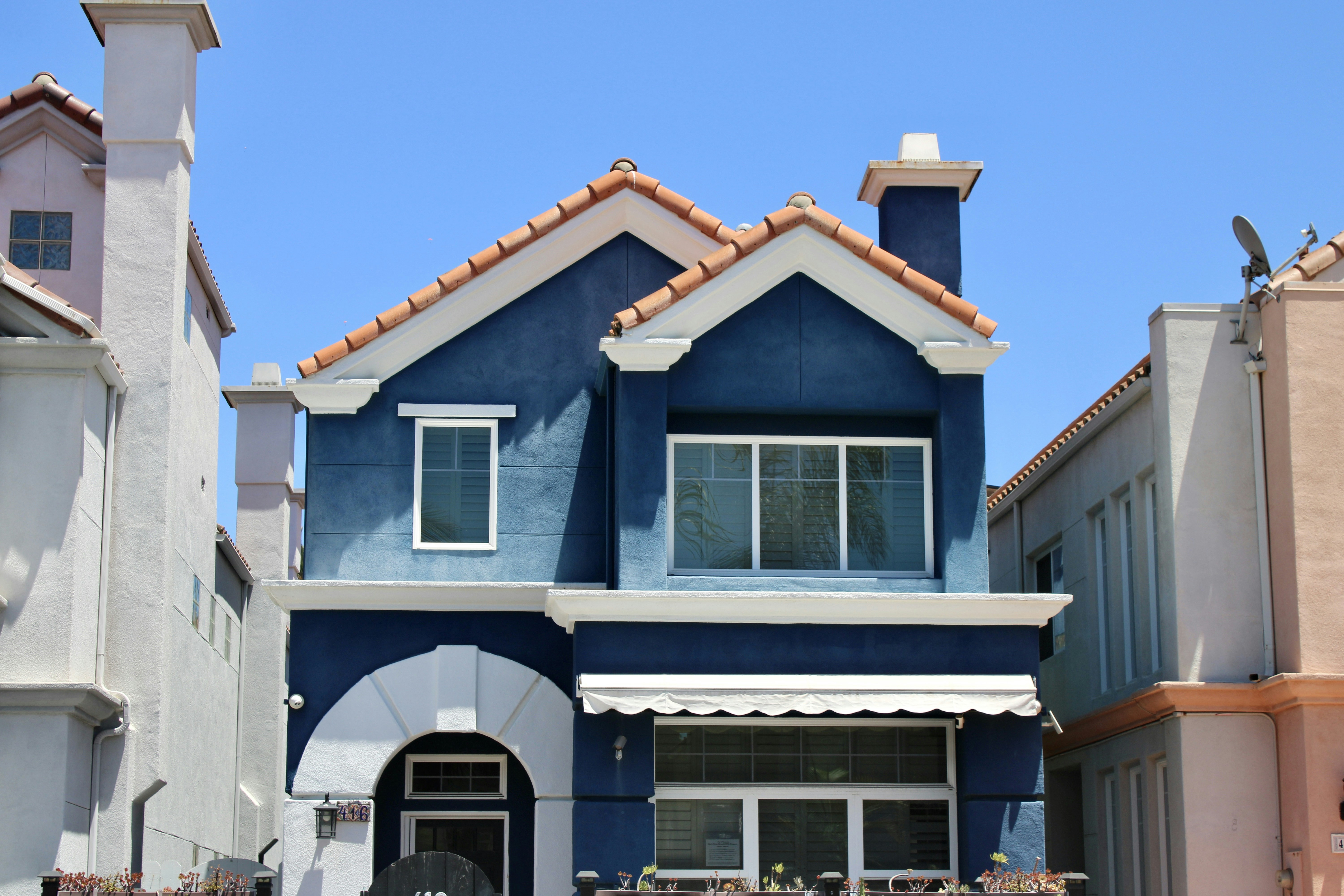 Vibrant blue house featuring a unique architectural design and decorative roof tiles, flanked by neighboring structures. The clear sky adds to the charm.