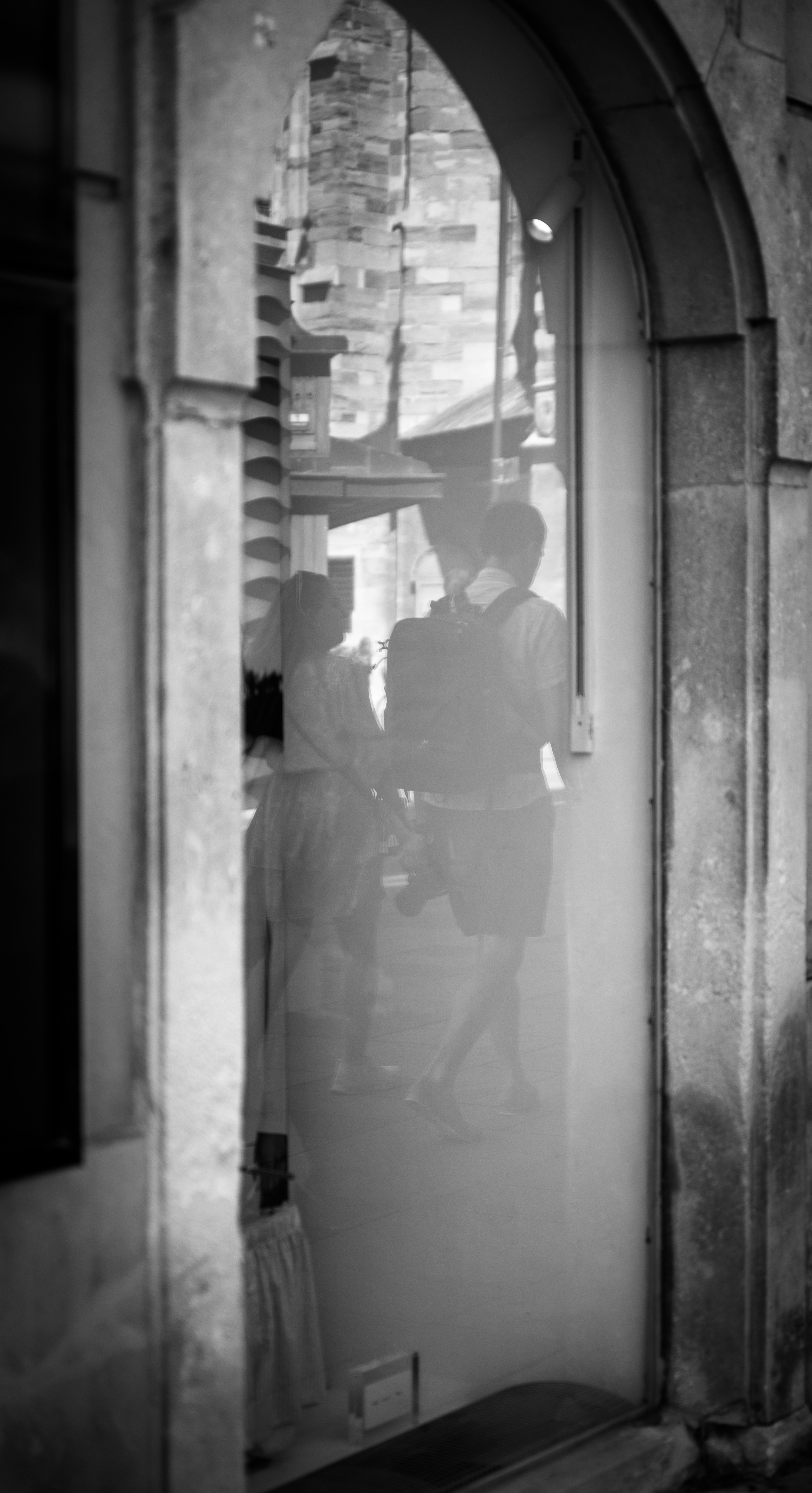 Two figures walking past a shop window, their reflections merging with the architecture behind them. A monochromatic scene that captures the essence of city life.