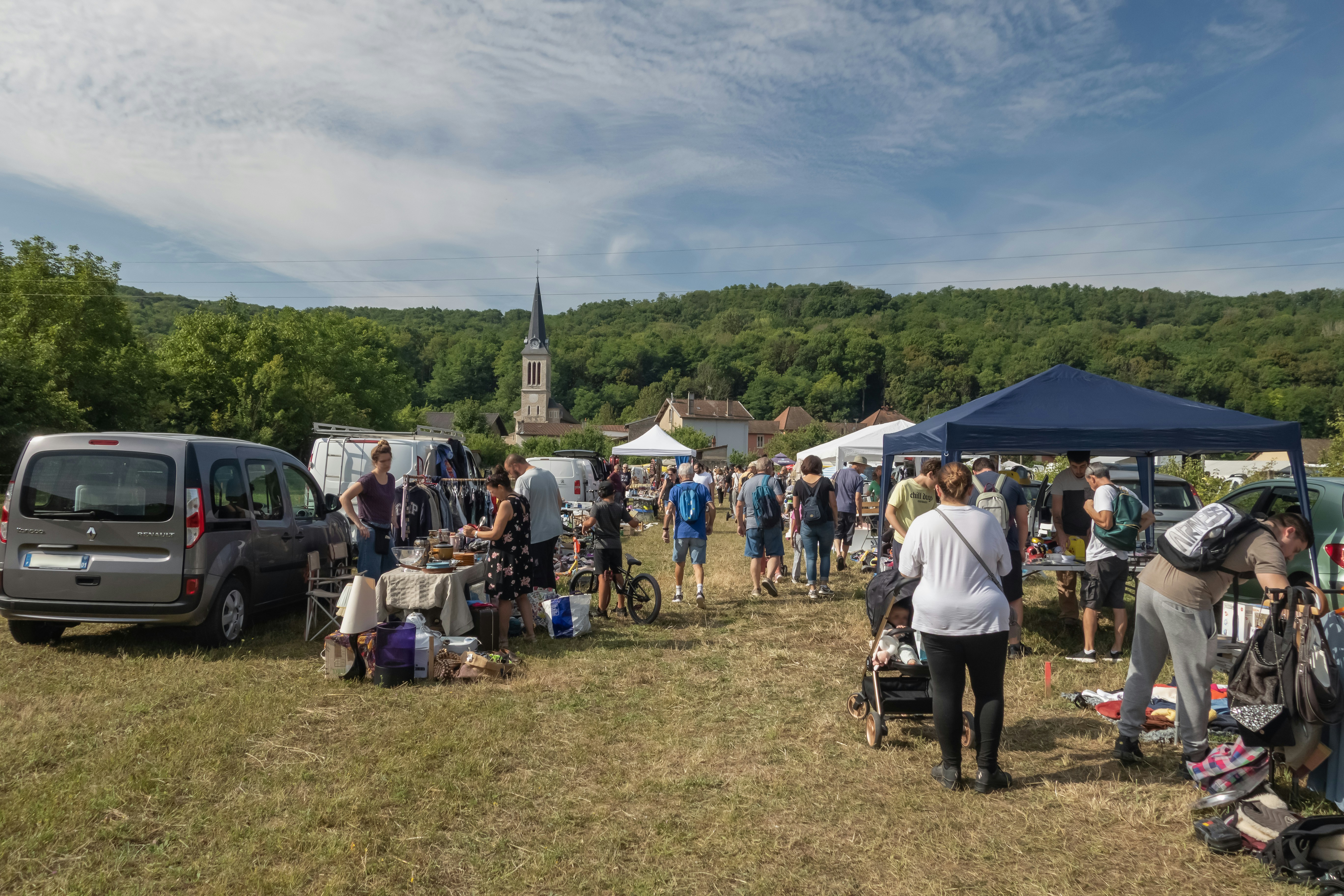 Le marché traditionnel du village de Bettant dans le Bugey en France. 🧺 Saveurs et savoir-faire au cœur du Bugey sous le ciel doux de cette fin de Juillet à Bettant, le marché s’éveille en couleurs et en parfums. Les paniers tressés racontent l’histoire des mains patientes, les étals débordent de produits du terroir, et les voix s’entrelacent dans une symphonie rurale. Ici, chaque sourire est une promesse de partage, chaque objet une trace vivante de l’artisanat français. Chaque étale est une chasse aux trésors pour tous. | A flea market is bustling outdoors on a sunny day.