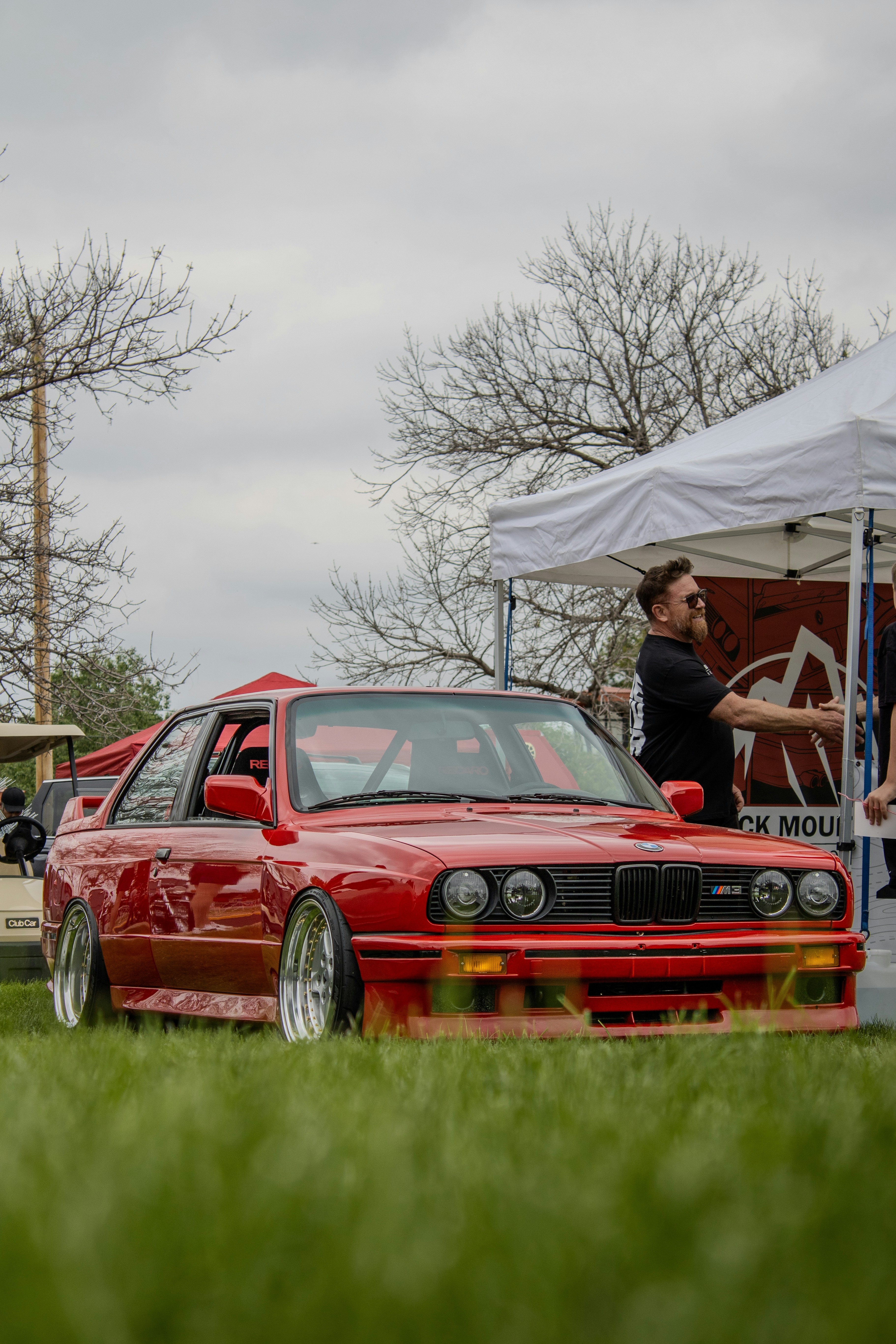 Modified red BMW parked on green grass with enthusiasts interacting in the background. The scene captures a vibrant car culture atmosphere.