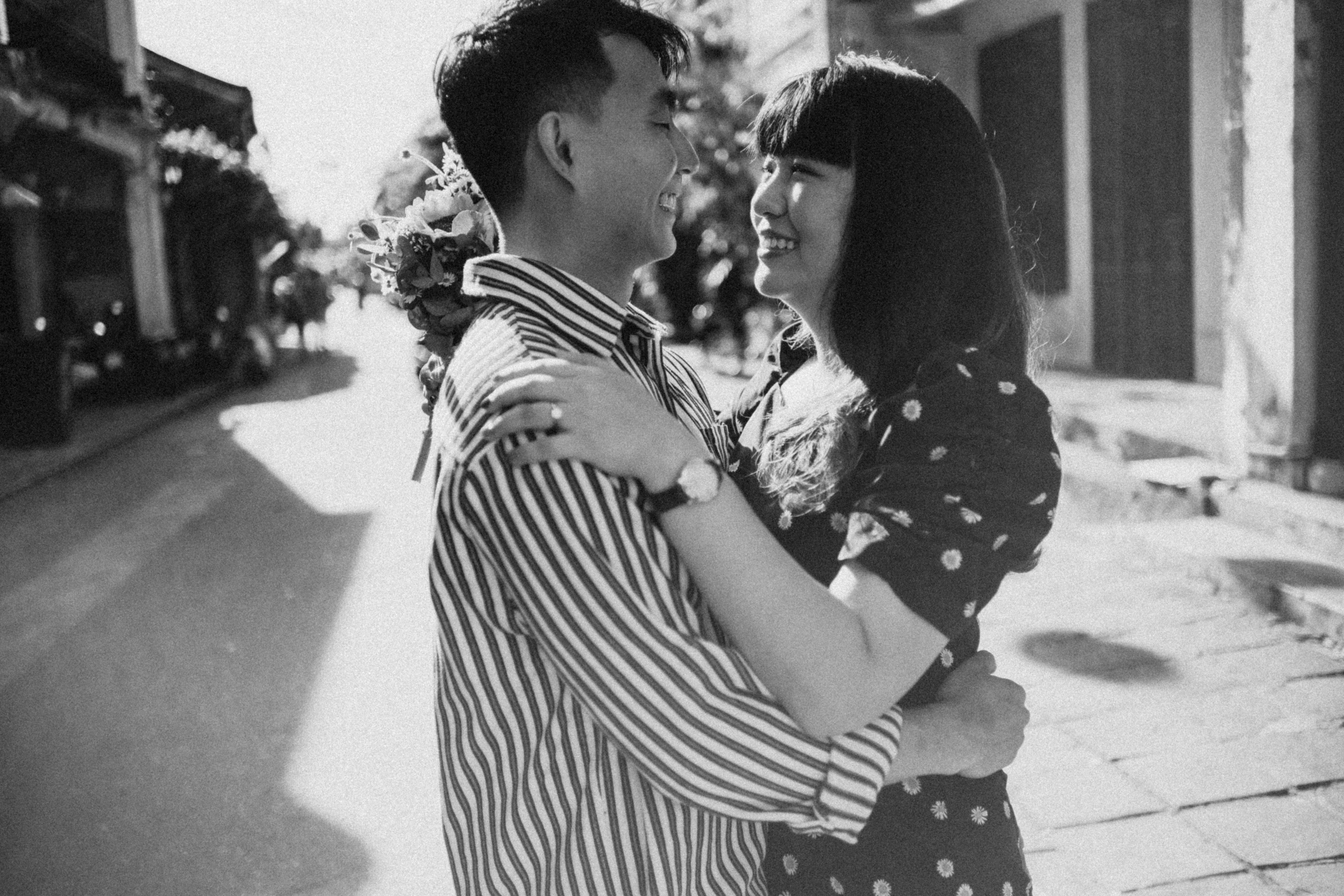 Couple embracing joyfully on a sunlit street, surrounded by charming architecture. Their expressions reflect a deep connection.