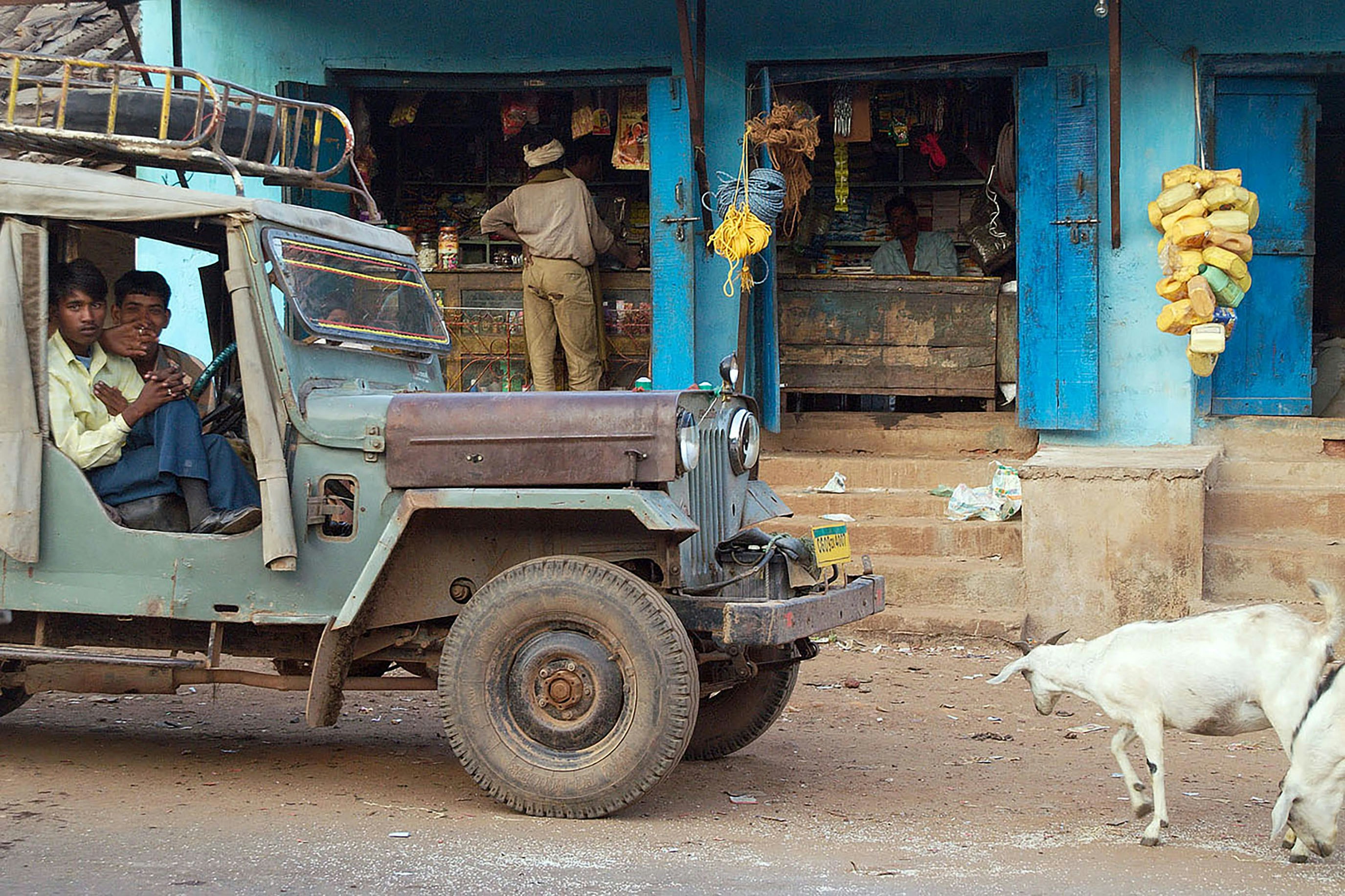 Two men seated in a vintage jeep while a goat wanders near a vibrant marketplace with blue storefronts.