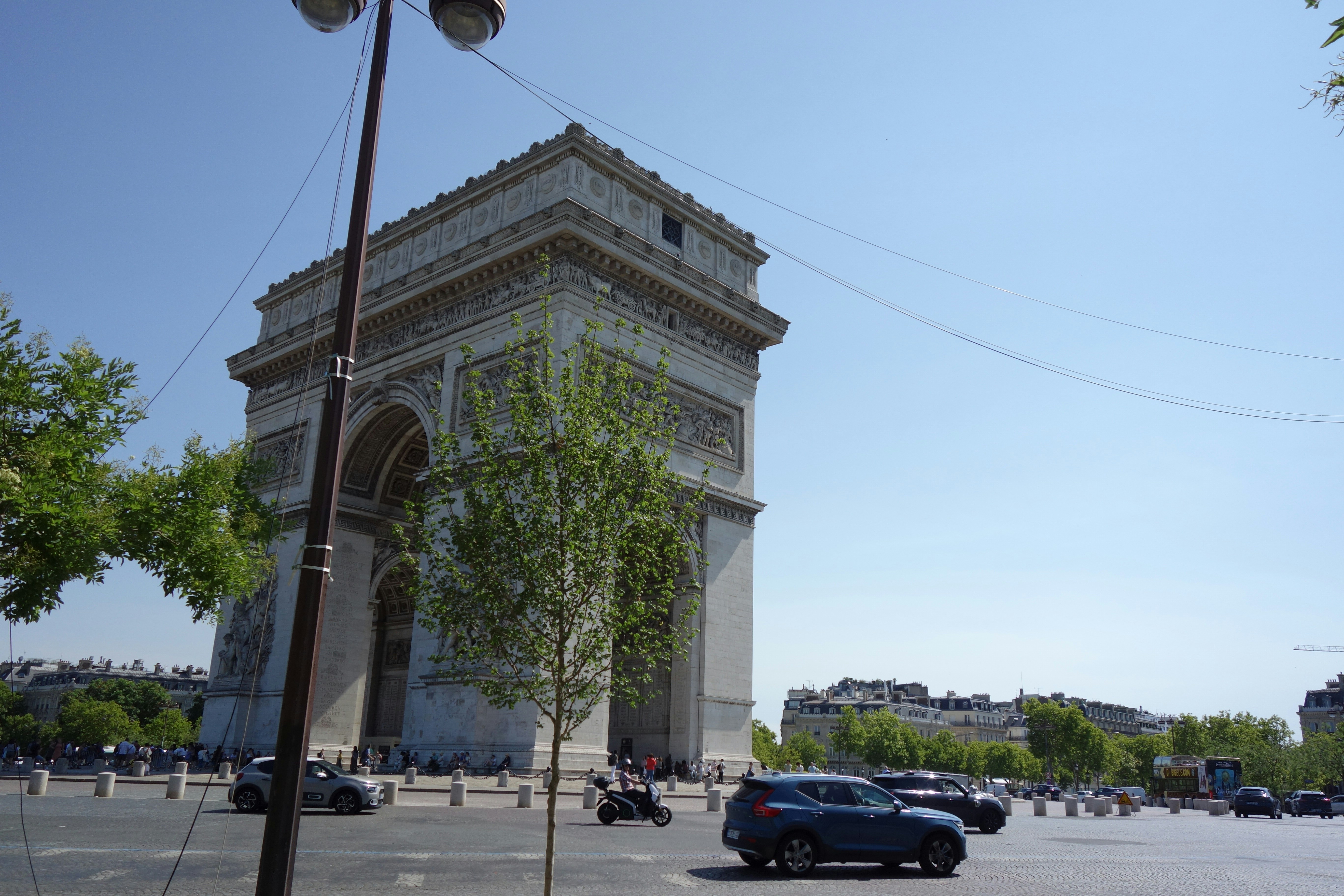 Arc de Triomphe stands majestically amidst bustling streets and greenery, showcasing its architectural splendor. The scene captures a harmonious blend of nature and urban life.