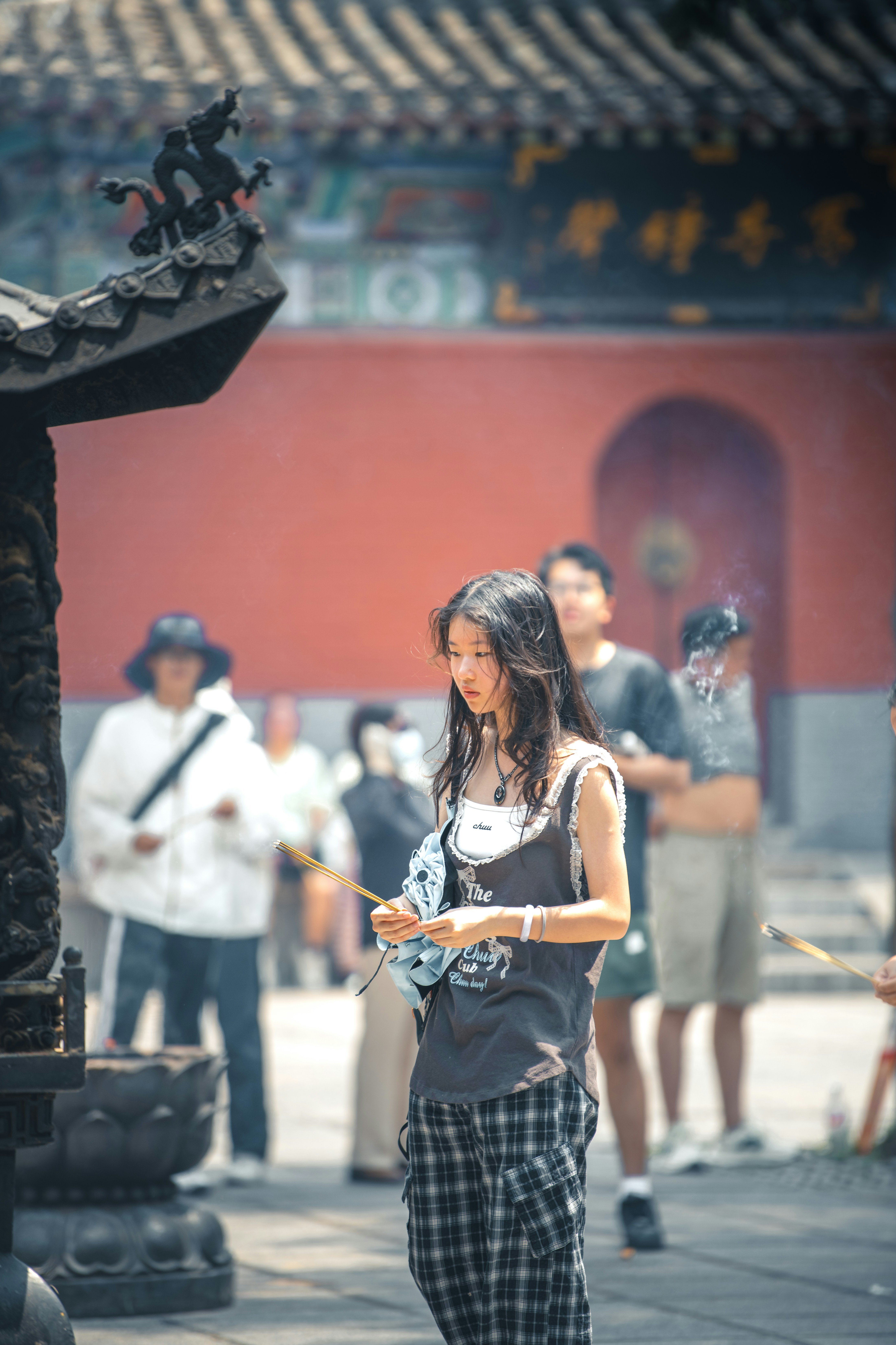 Young woman holding incense sticks in a temple courtyard, surrounded by visitors engaging in prayer. Smoke wafts gently in the air.