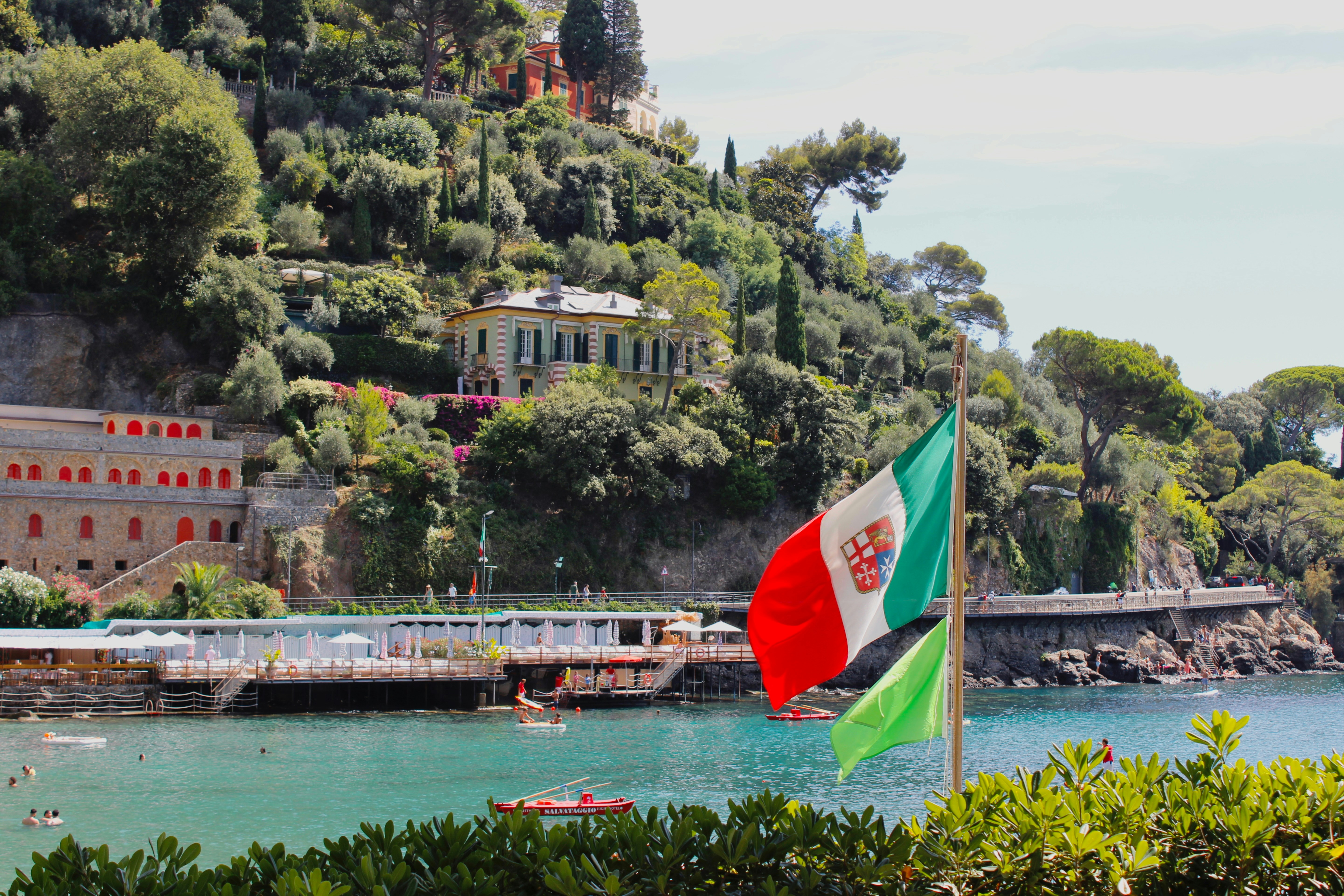 Italian flag waving proudly in the foreground, framed by lush greenery and colorful coastal villas. The tranquil waters reflect the vibrant scenery.