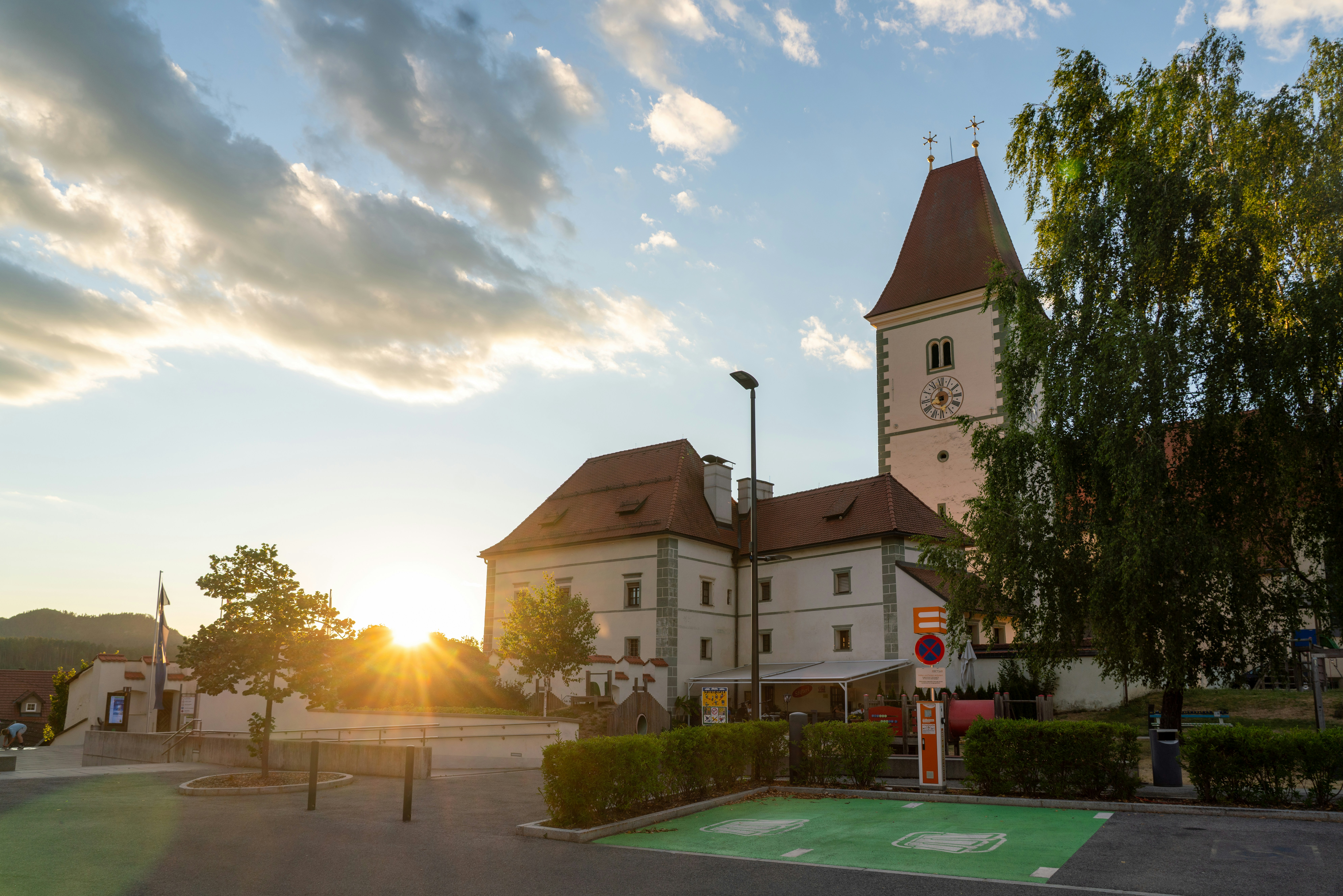 Historic building with a clock tower illuminated by the setting sun, surrounded by lush greenery and urban elements.
