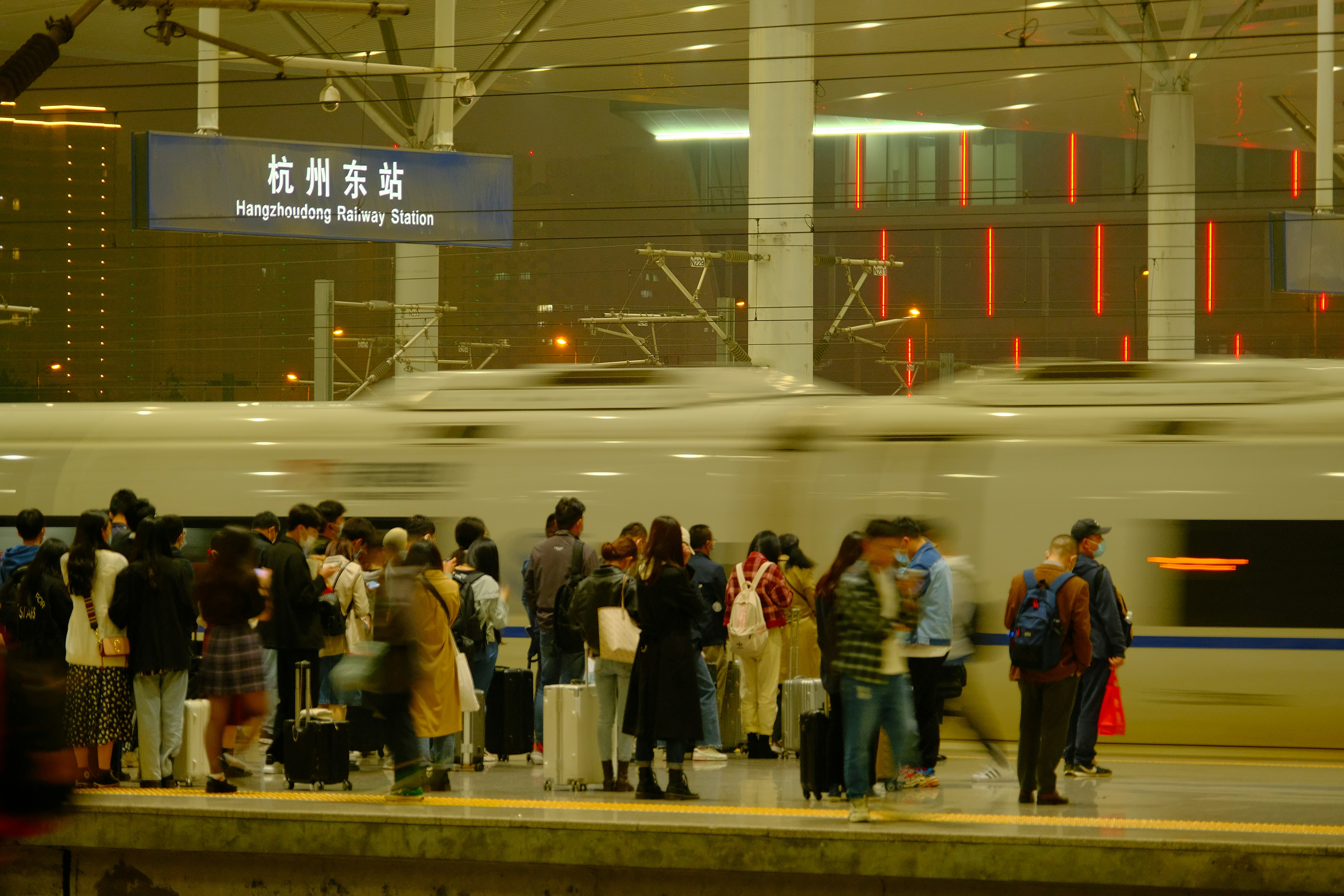 Crowd of travelers waiting at Hangzhou Railway Station as a high-speed train rushes by, capturing the essence of modern travel in a bustling city.