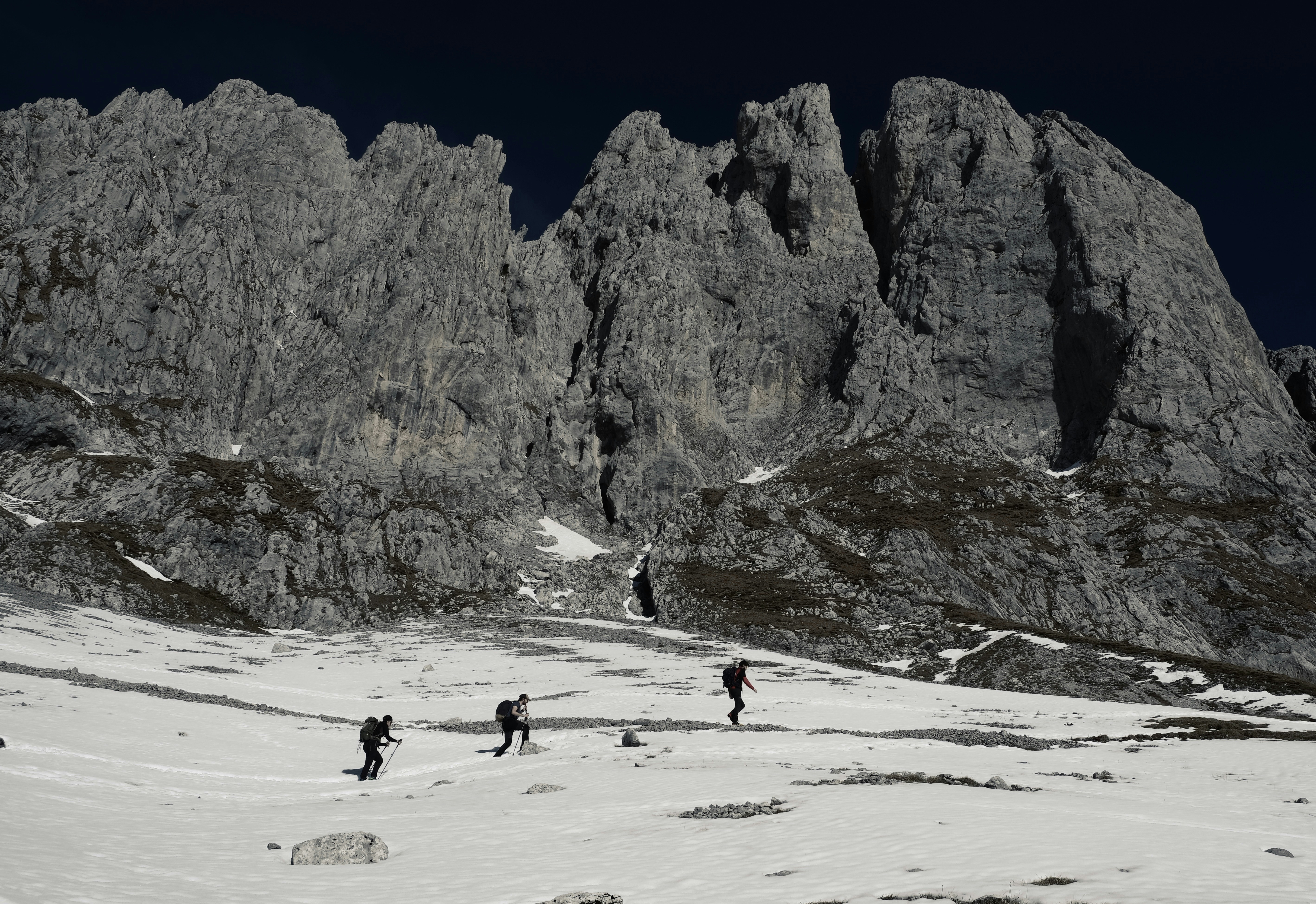 Trekking ai piedi della Presolana | Hikers ascend a snowy slope towards rocky mountains.