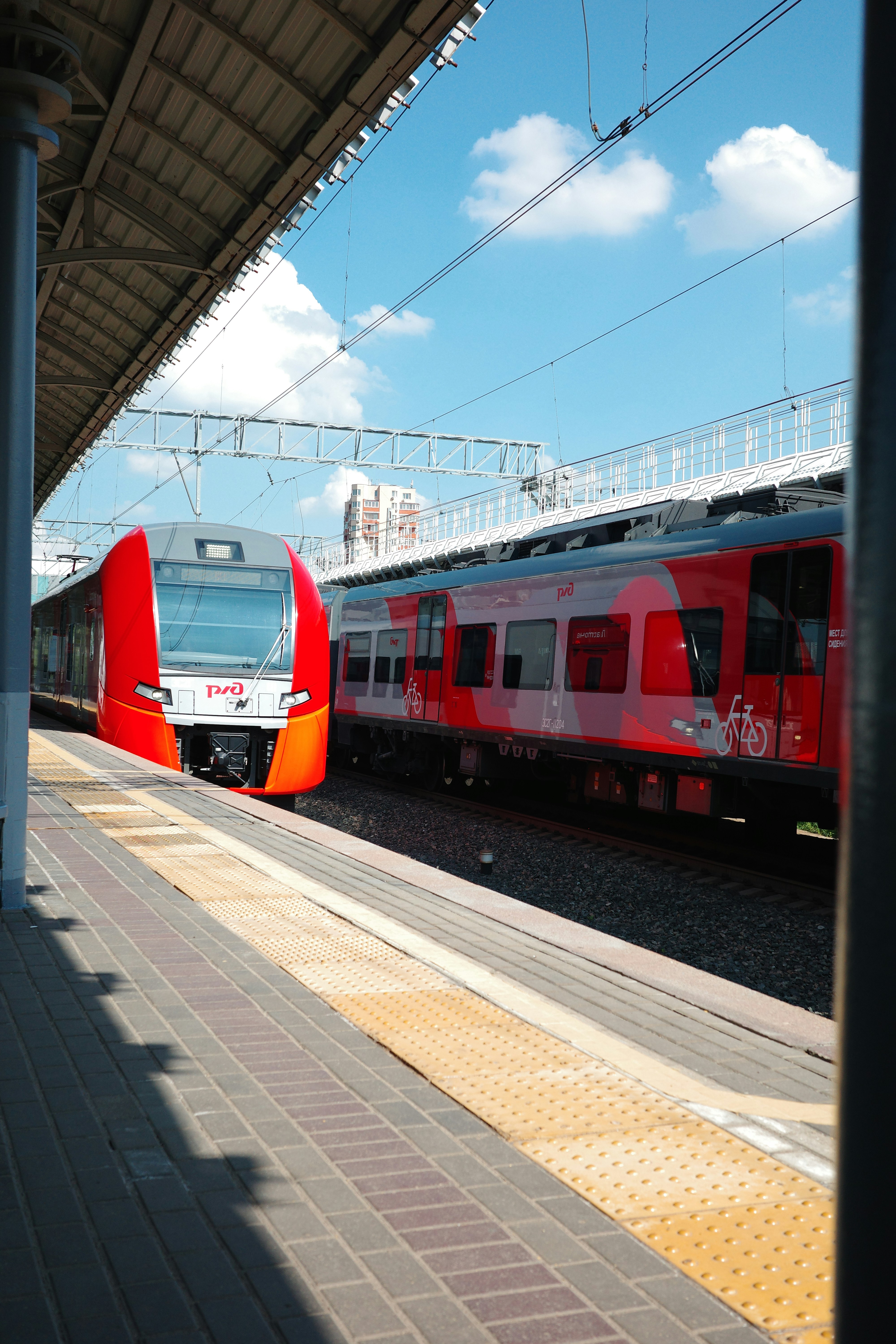 Two trains at a bustling station, one sleek red and the other adorned with bicycle graphics, under a bright blue sky.