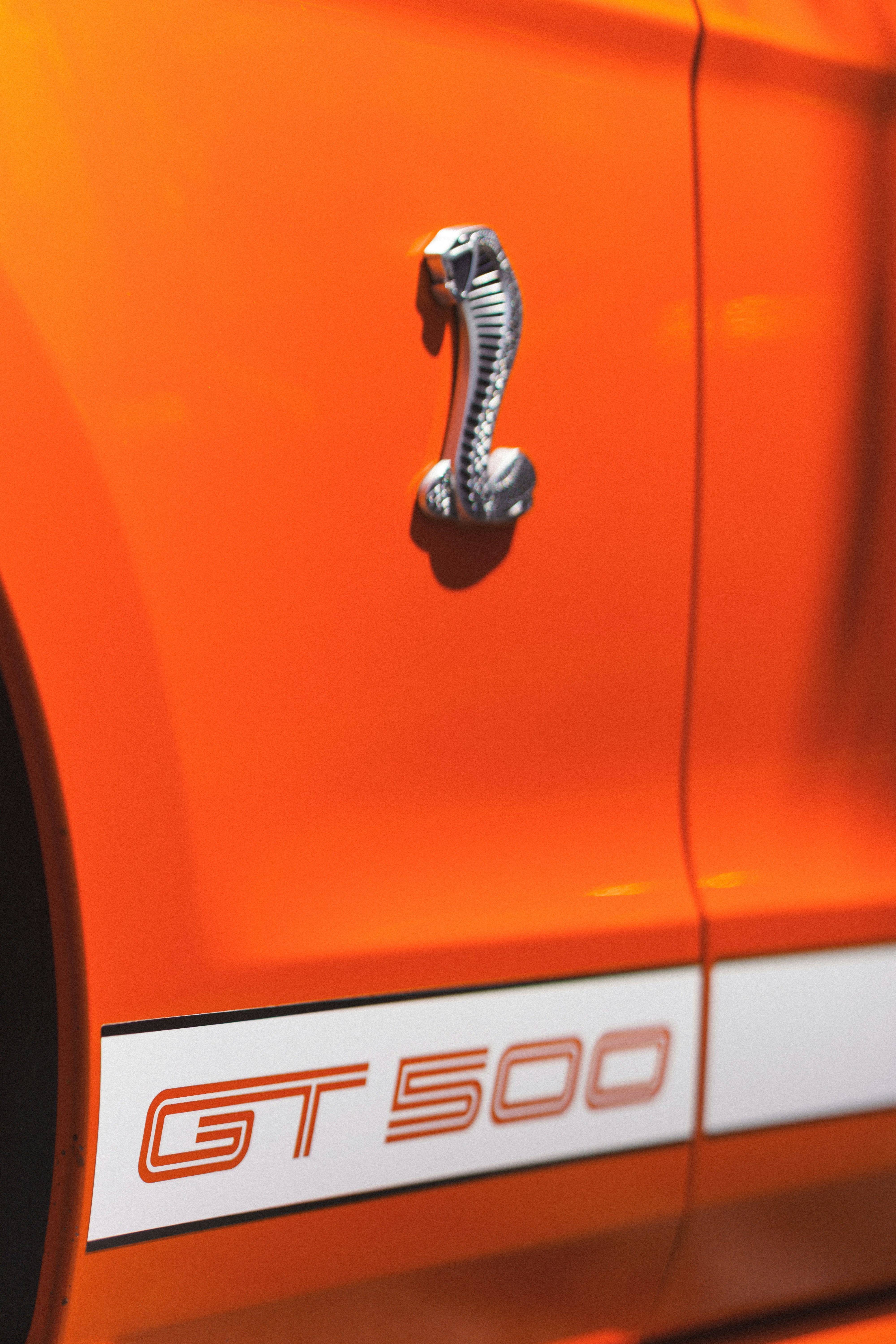 Close-up of a Shelby Cobra emblem on an orange GT500, showcasing the vehicle's distinctive branding and vibrant color.