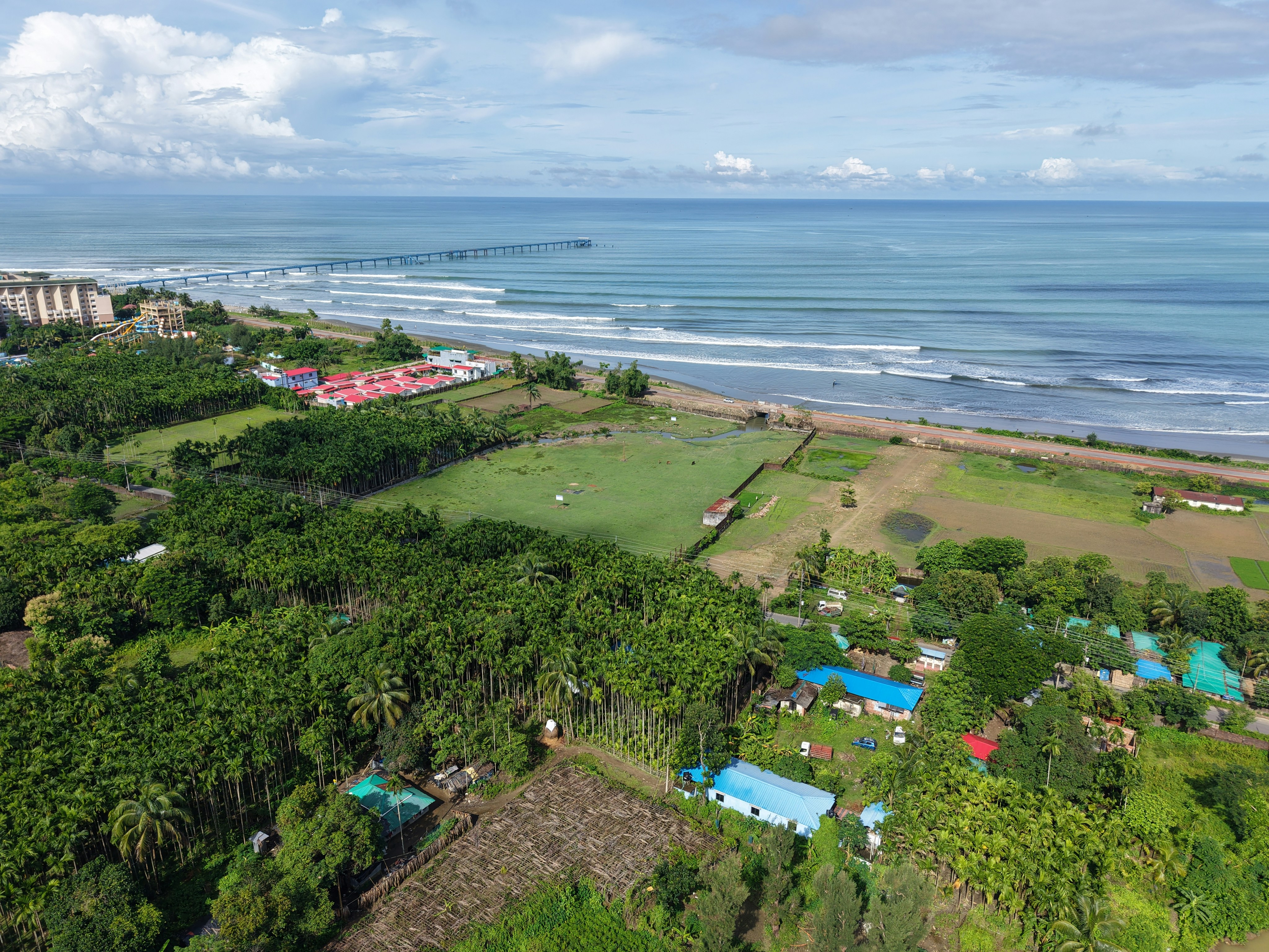 Aerial View of Inani Beach and Palm Groves, Cox’s Bazar This breathtaking aerial shot captures the natural beauty of Inani Beach, located in Cox’s Bazar, Bangladesh — the world’s longest natural sea beach. Taken with a DJI Air 3S, the image showcases lush palm groves, coastal farmland, and the serene blue waters of the Bay of Bengal. The long pier stretching into the ocean adds depth to this tropical paradise. Ideal for themes around travel, nature, tropical destinations, and sustainable living. | Aerial view of a lush, coastal landscape.