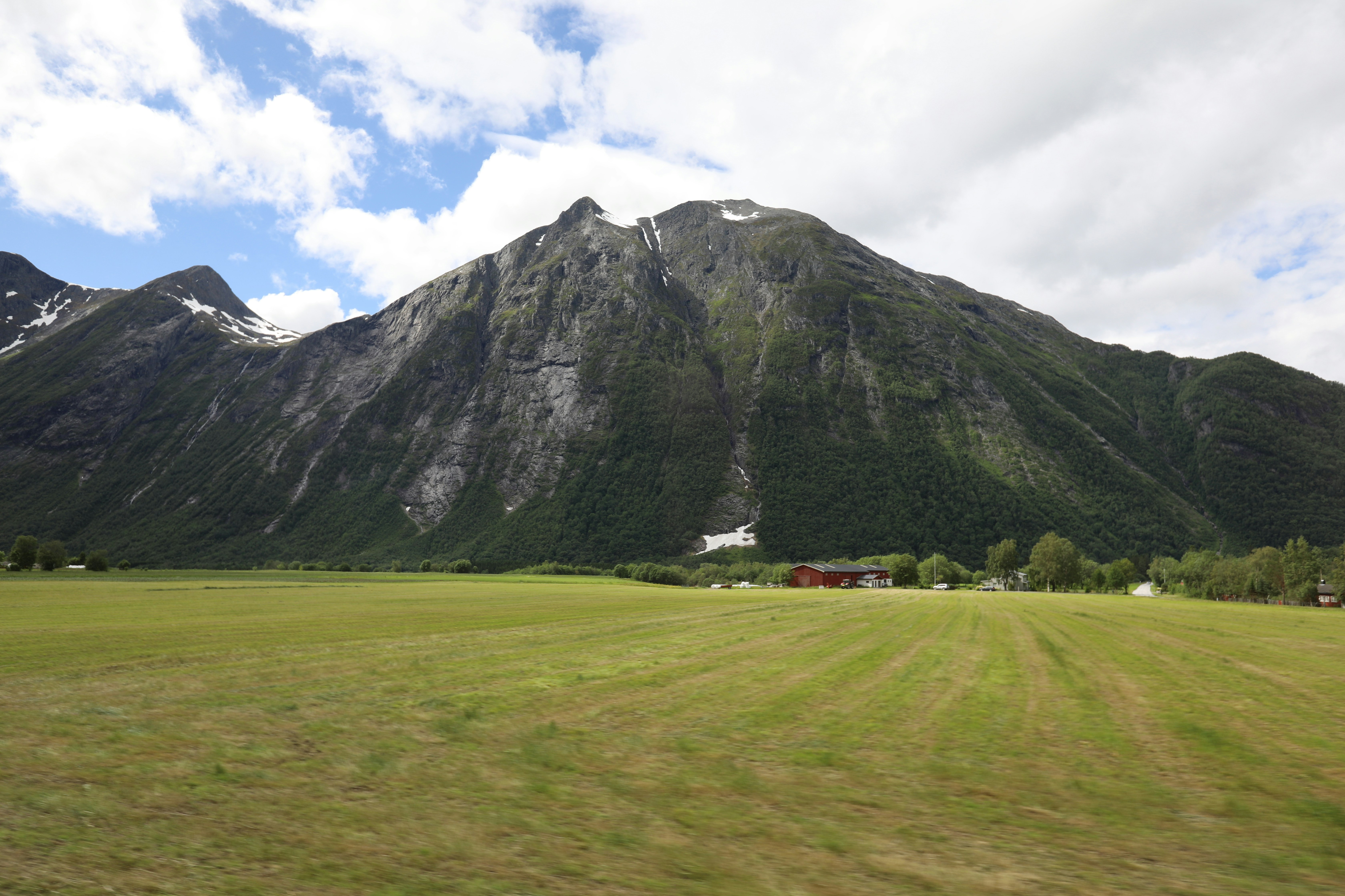 Lush green valley framed by towering mountains under a partly cloudy sky. A few farm structures dot the landscape.