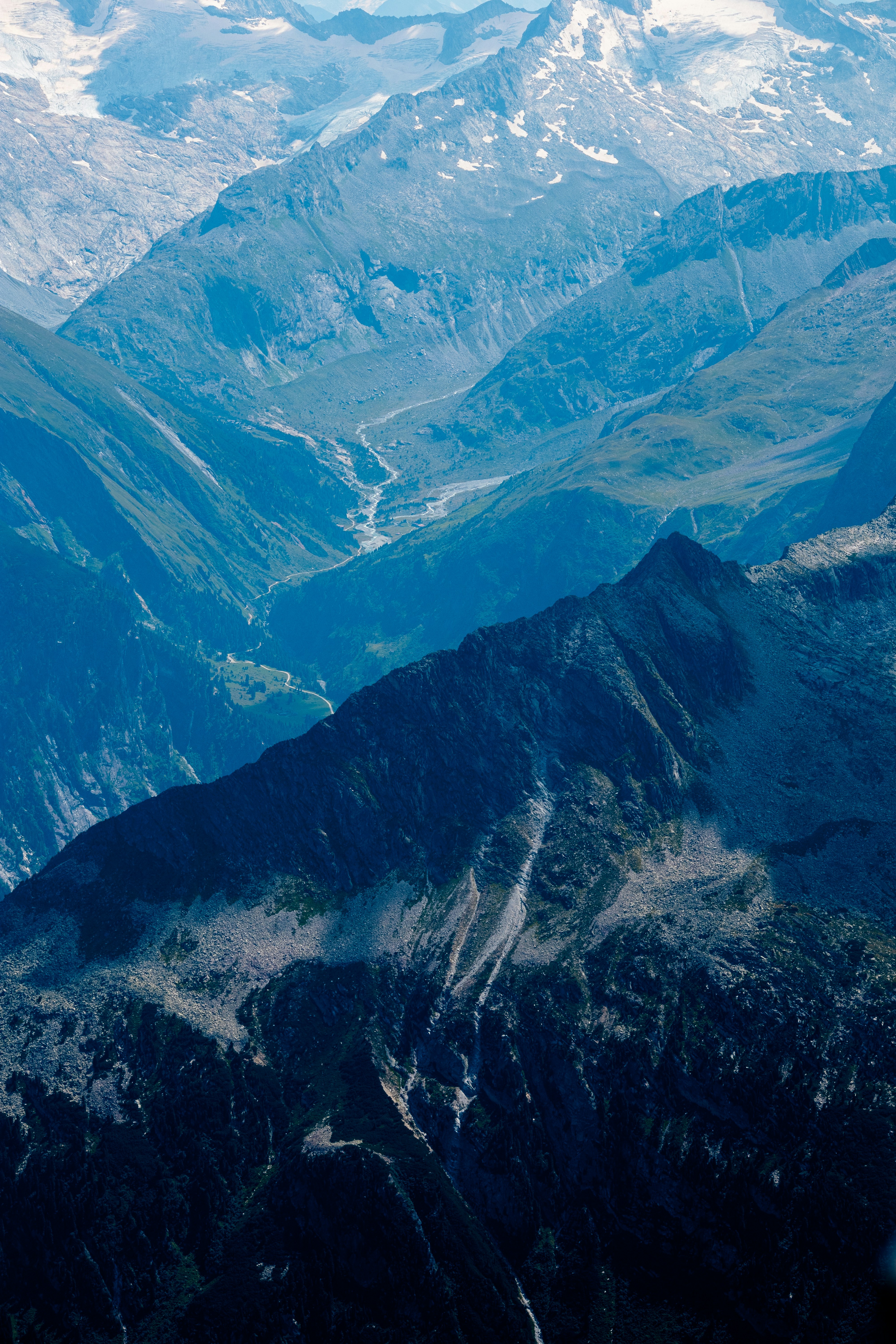 Dramatic mountain ranges and valleys showcase the rugged beauty of the Alps, revealing intricate textures and shadows. A serene river winds through the lush landscape below.