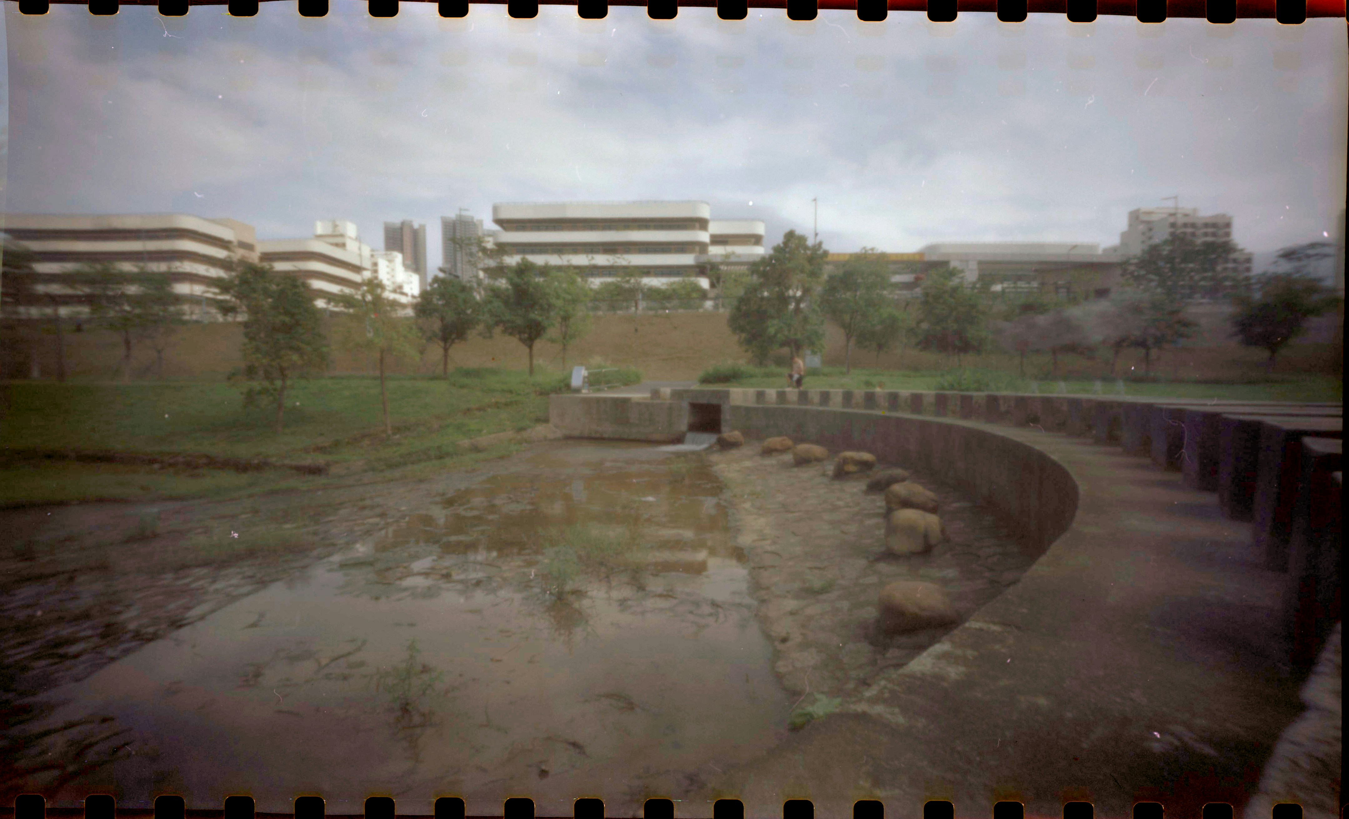 A serene view of a waterlogged park area, featuring modern buildings in the background and a curved stone structure leading into the water.