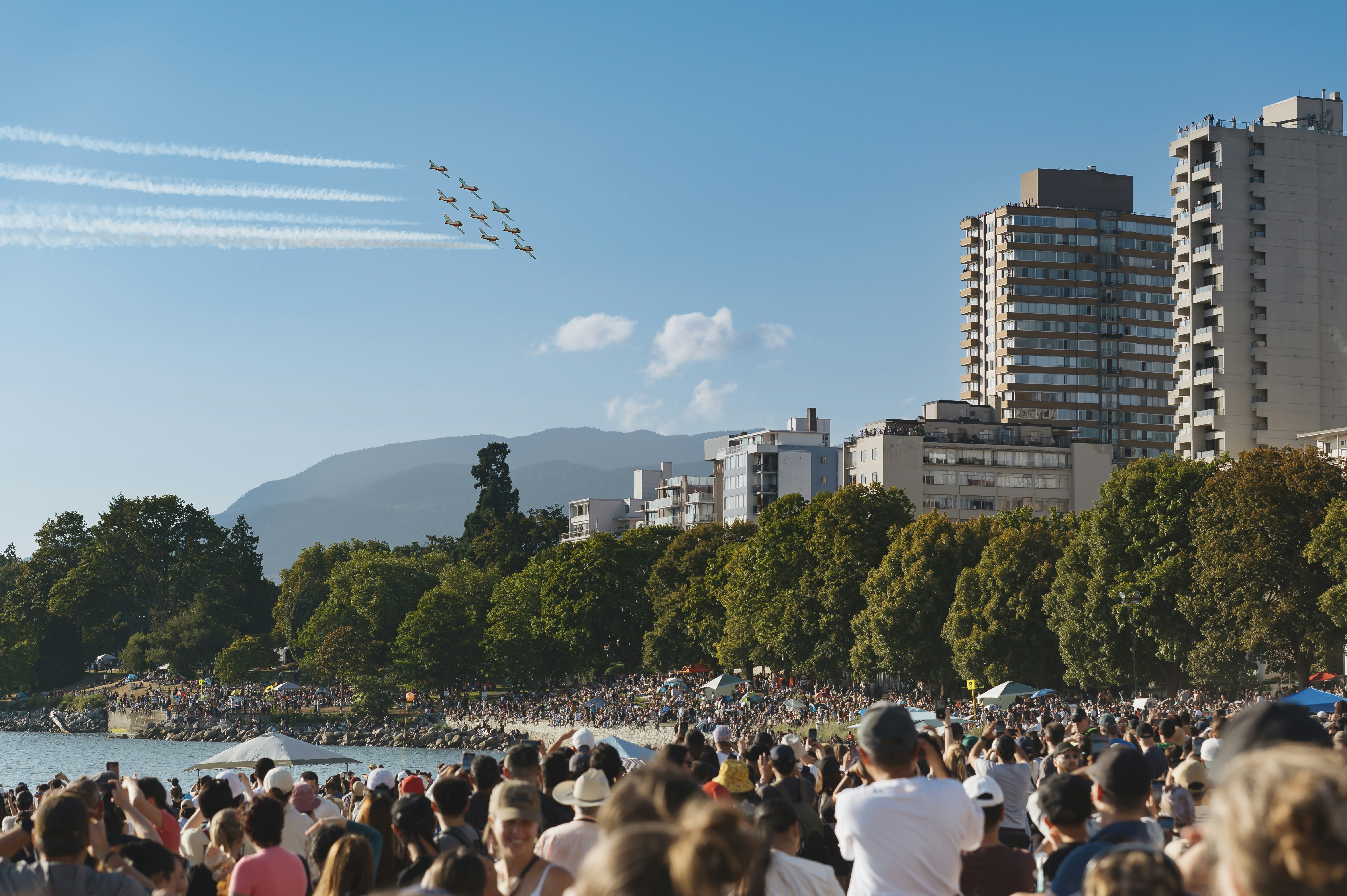 The Canadian Snowbirds flying over English Bay in Vancouver for the 2025 Honda Celebration of Lights | Airplanes fly above a crowd-filled city beach.