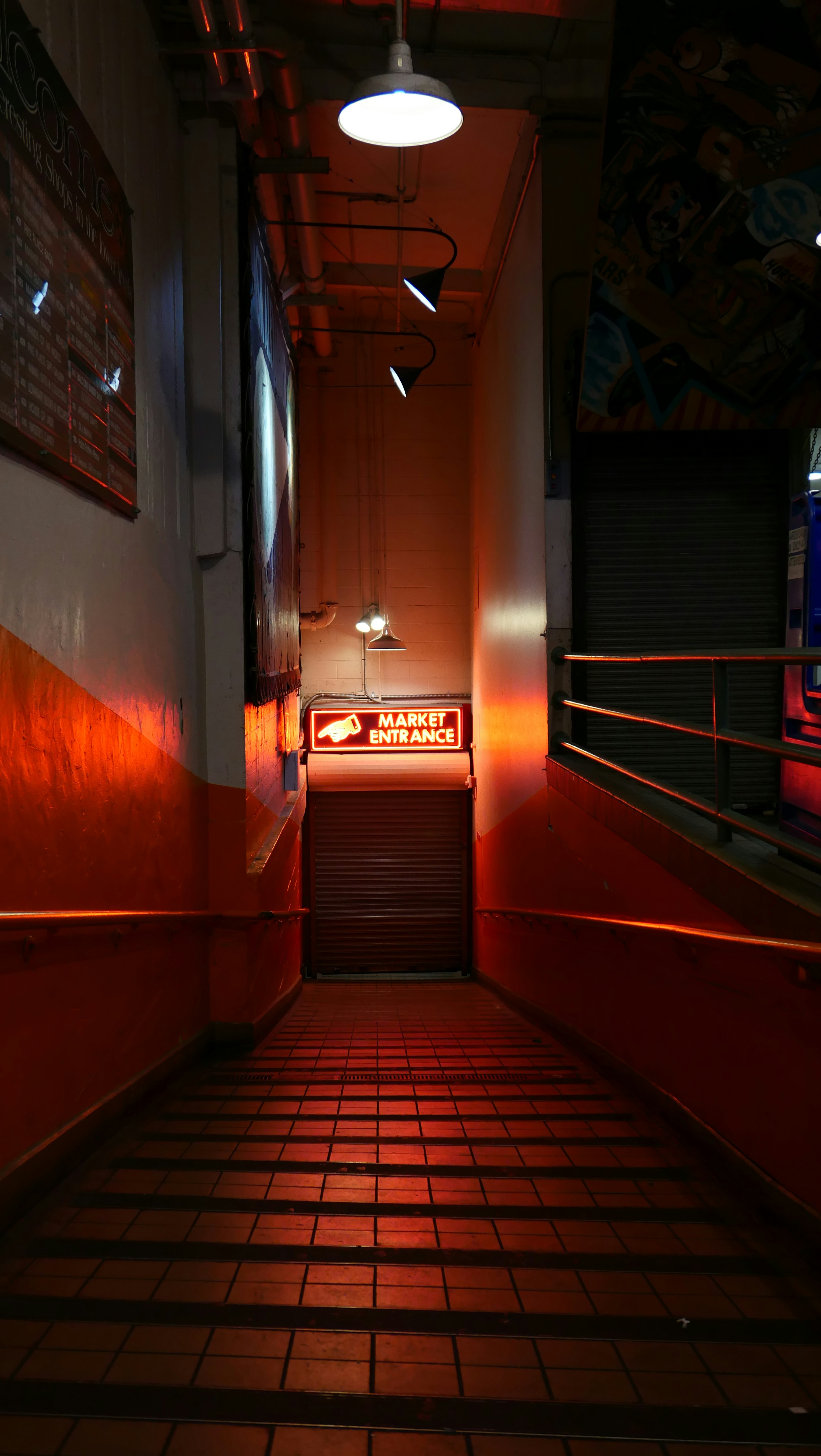 Neon sign reading 'MARKET ENTRANCE' illuminates a descending staircase, framed by warm orange walls and soft overhead lighting.