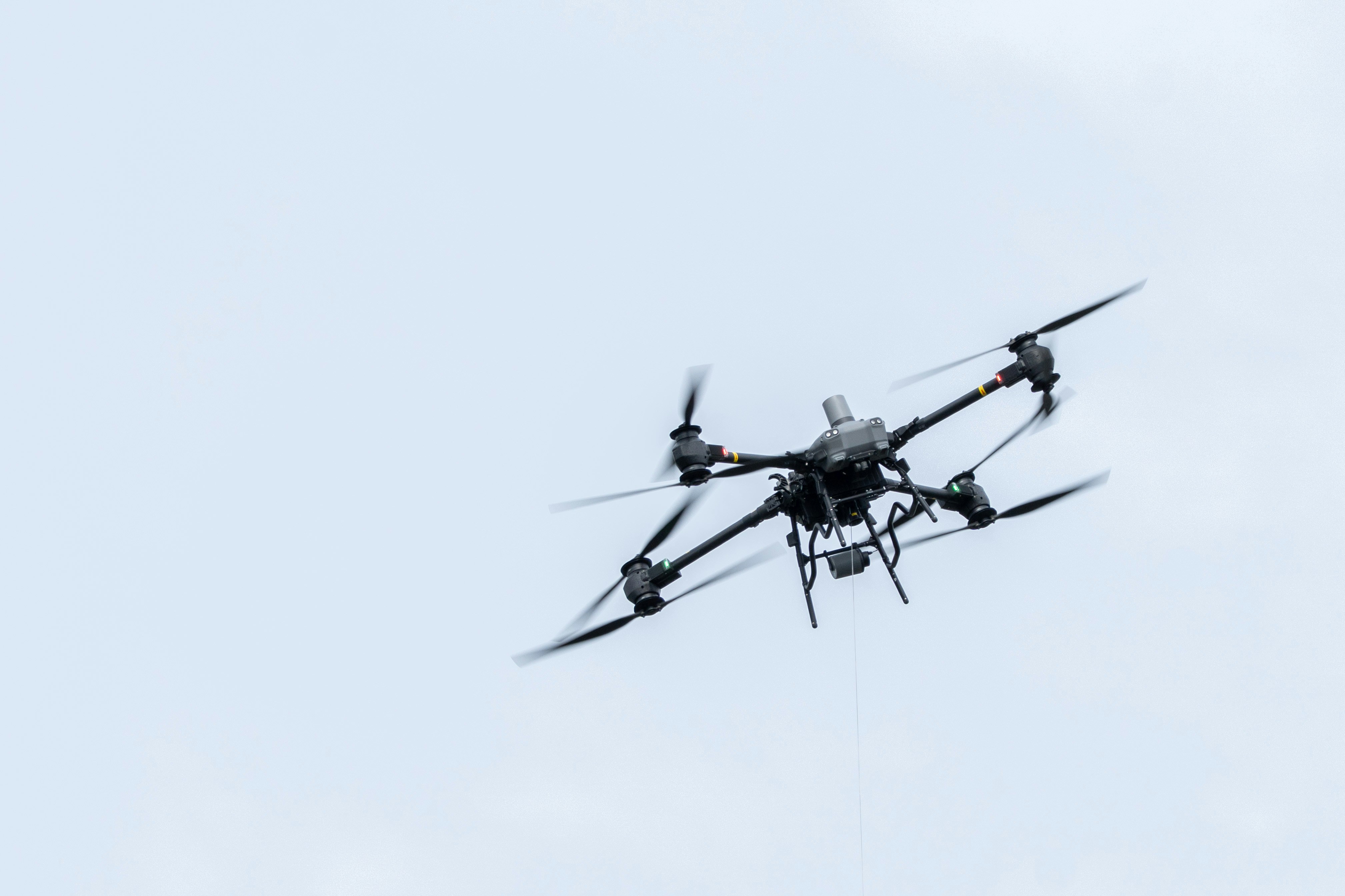 A close-up view of a heavy-lift drone in mid-flight against a pale sky, equipped with multiple rotors and a suspended cable—highlighting the power and precision of aerial delivery technology. | A drone flies against a cloudy sky.