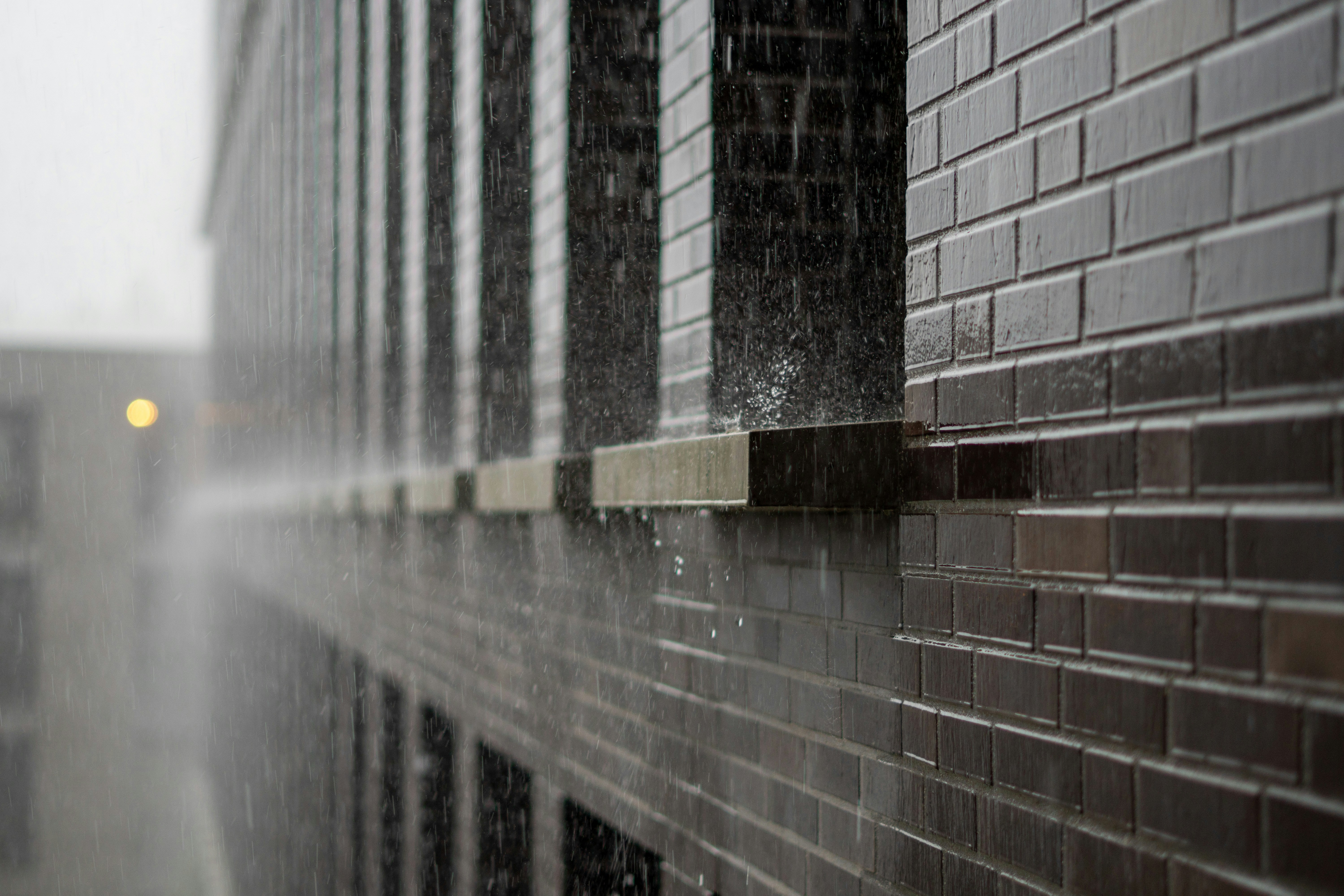 Heavy rain pours down a dark brick building, with water cascading off ledges and streaming down the walls—capturing the raw power of a storm against the stark geometry of urban architecture. | Rain falls on a brick building.