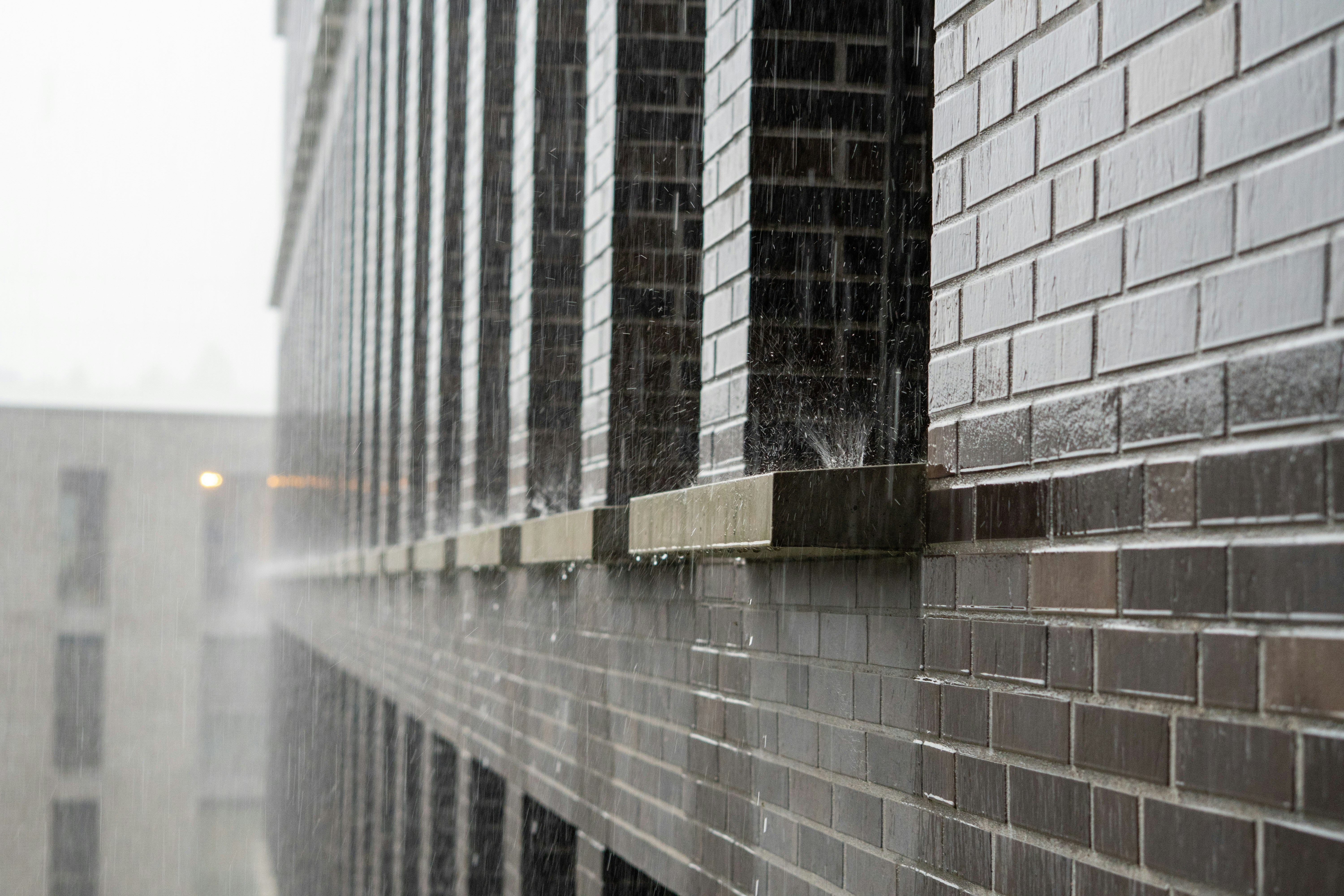 Torrential rain lashes against the dark brick facade of a modern building, with water splashing from ledges and cascading down the walls—capturing the intensity of a downpour in an urban setting.