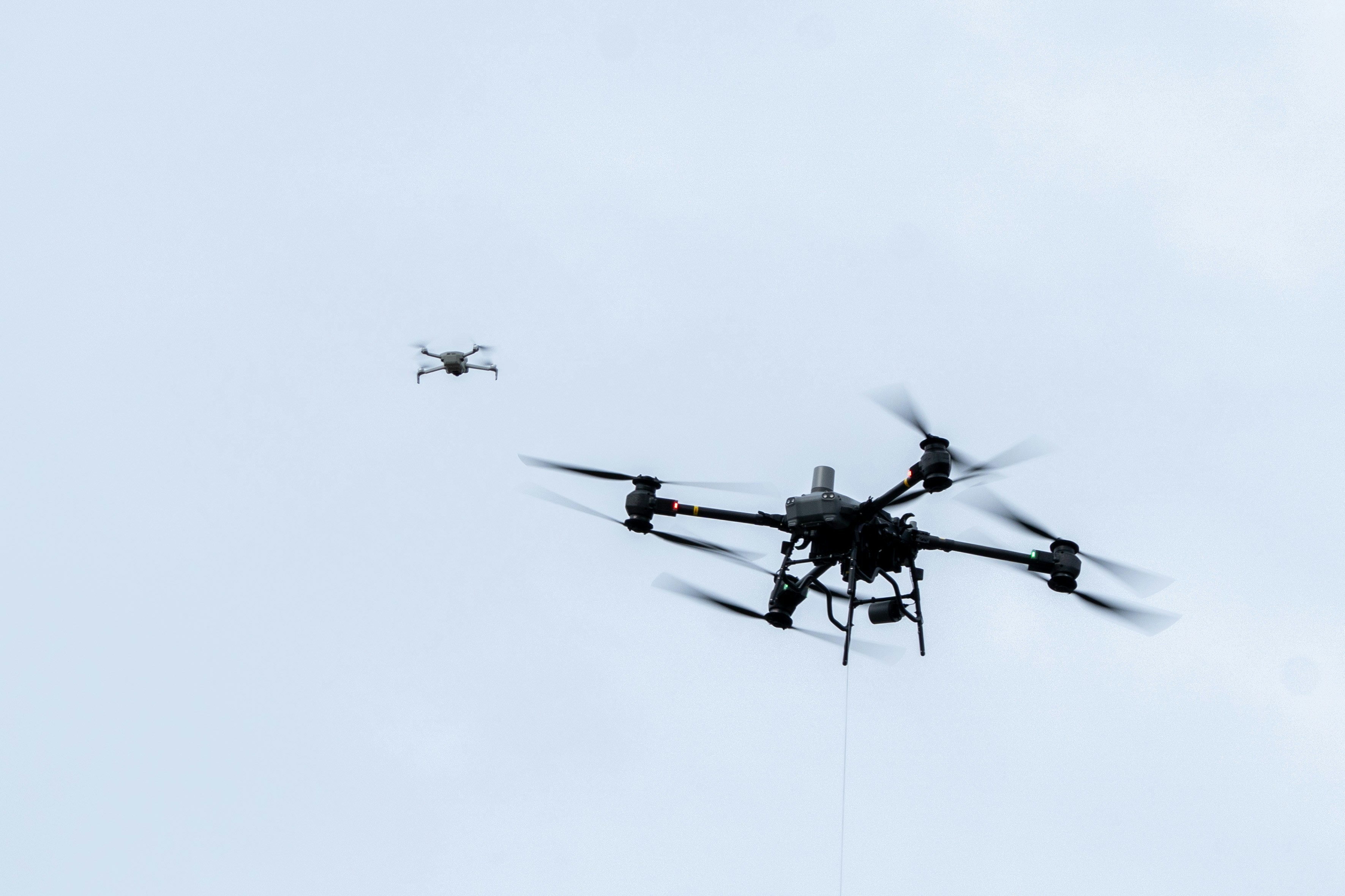 Two drones soar through a pale sky—one large industrial drone actively transporting solar panels, while a smaller quadcopter observes nearby. A visual contrast of scale, purpose, and the evolving capabilities of aerial technology. | Drones are flying in a cloudy, blue sky.