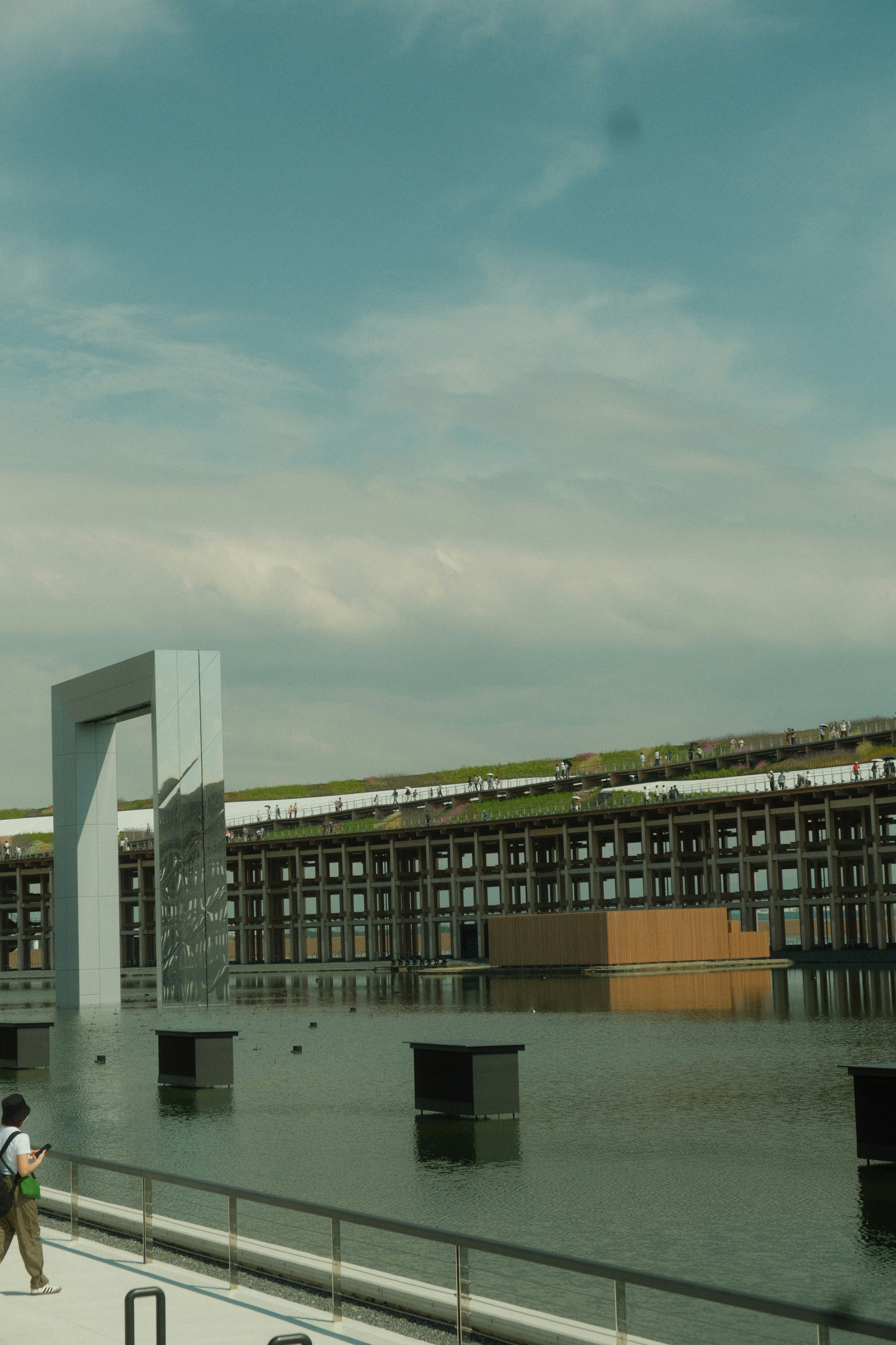 A sleek, mirrored arch stands beside a serene water body, surrounded by modern architecture and lush greenery. The scene captures a harmonious blend of nature and contemporary design.