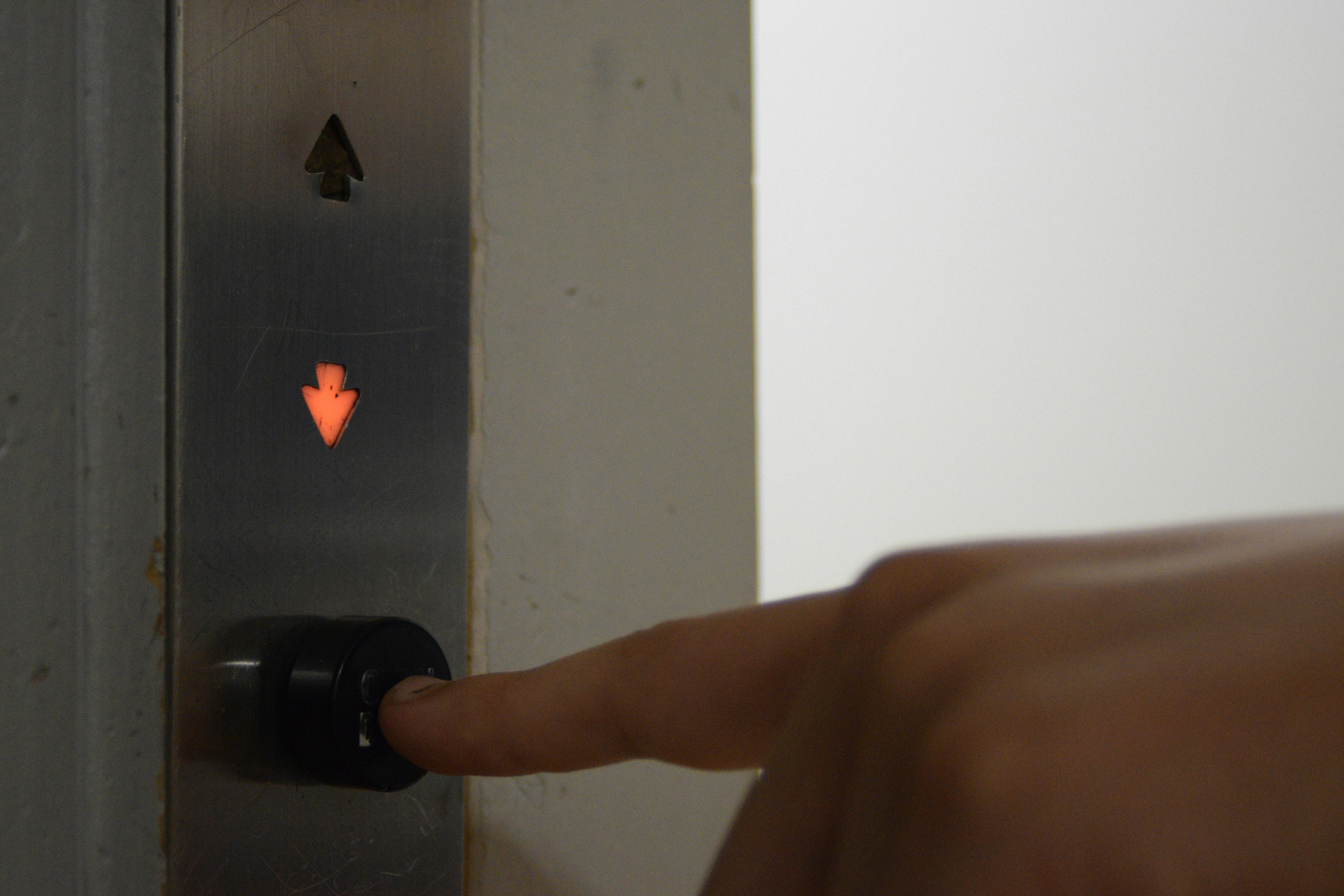 A hand poised to press the down button on an elevator panel, with the illuminated arrow indicating the direction. The metallic surface reflects a hint of the surrounding environment.