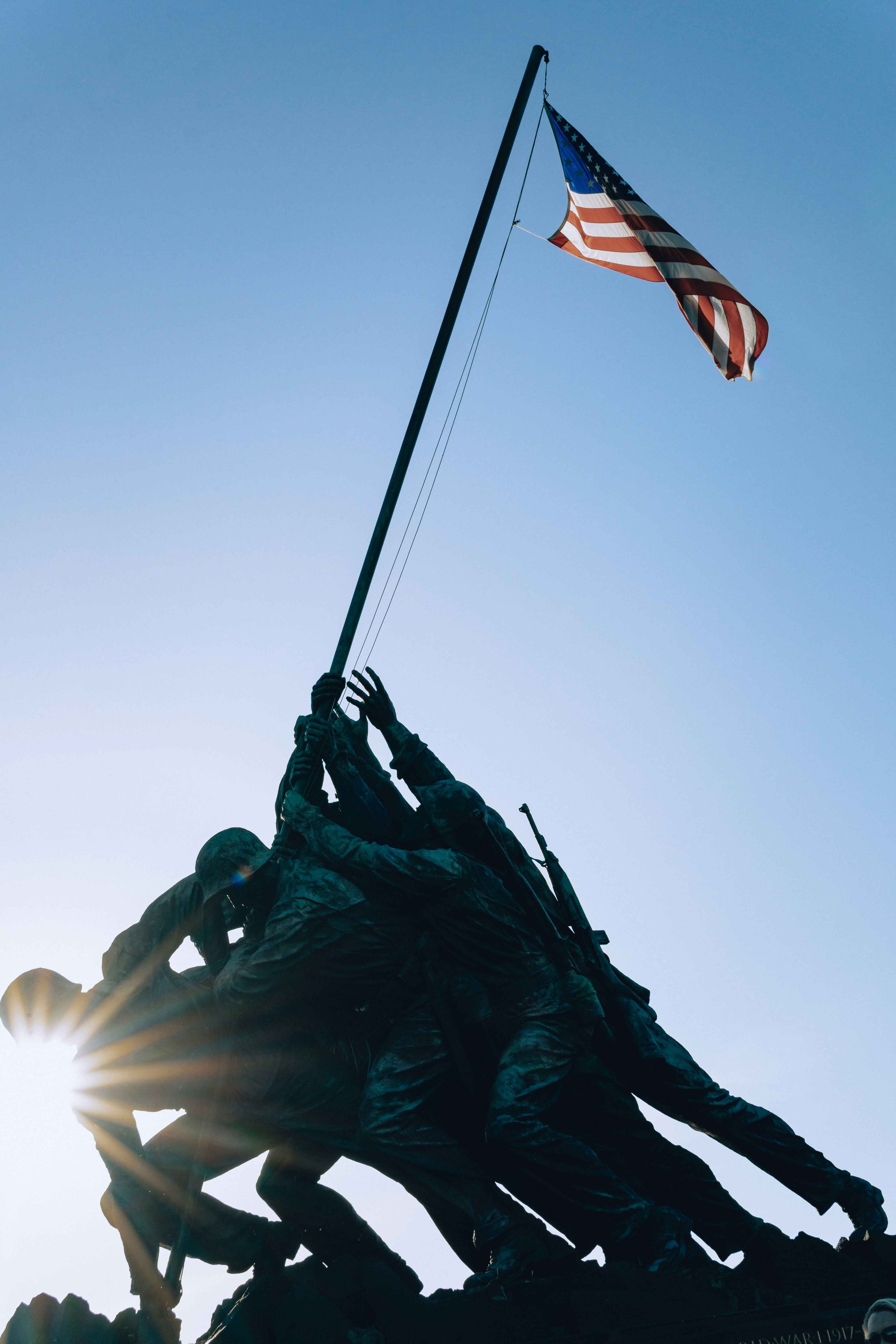 Bronze statue depicting soldiers raising the American flag against a clear blue sky, with sunlight creating a dramatic halo effect.