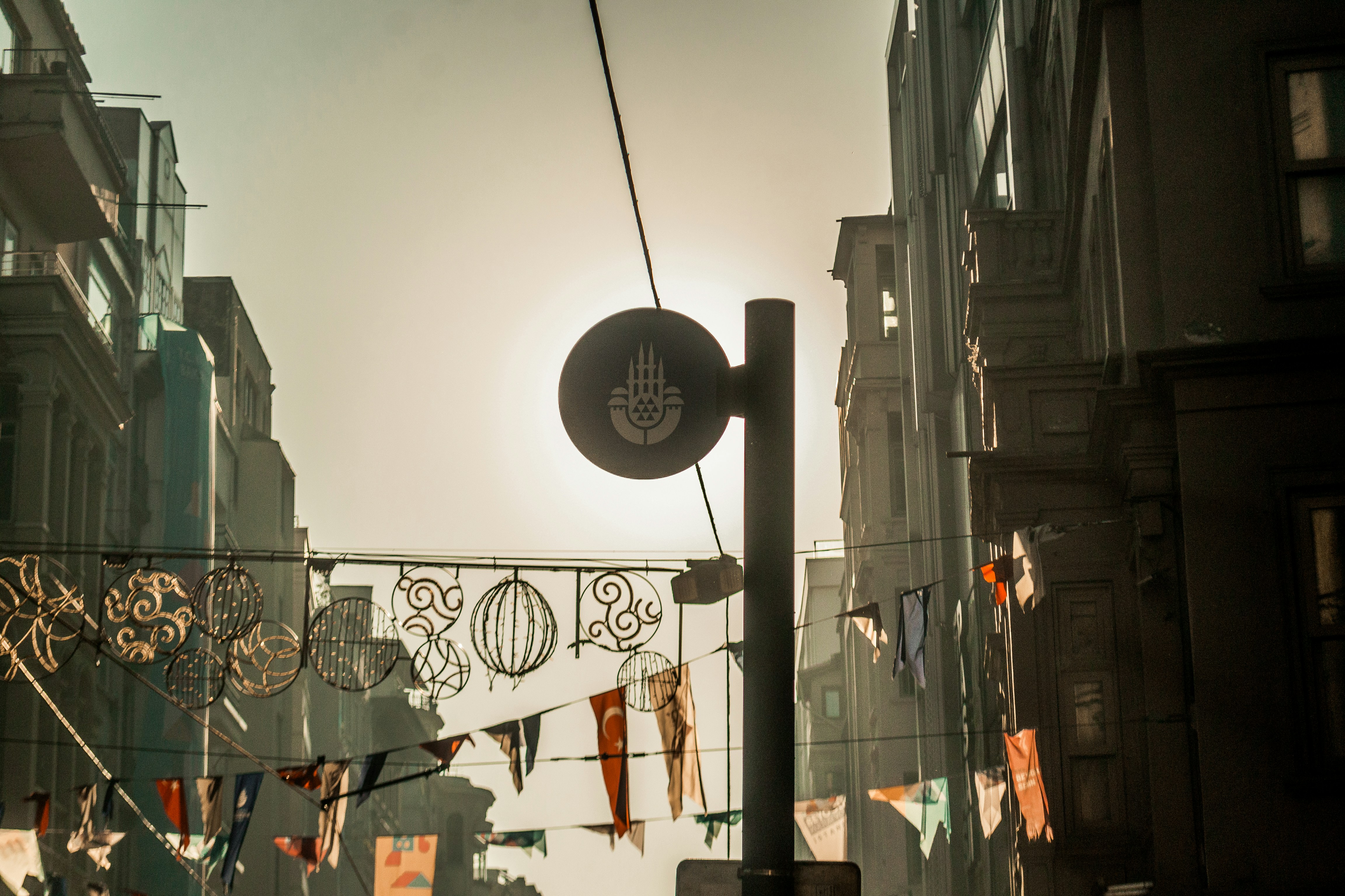 Sunlight aligns perfectly with a municipal sign on a narrow Istanbul street, creating a radiant halo effect. Draped flags and decorative street lights frame the scene, blending old-world charm with modern urban energy in the heart of the city. | Old-town street, decorated with flags and lights.