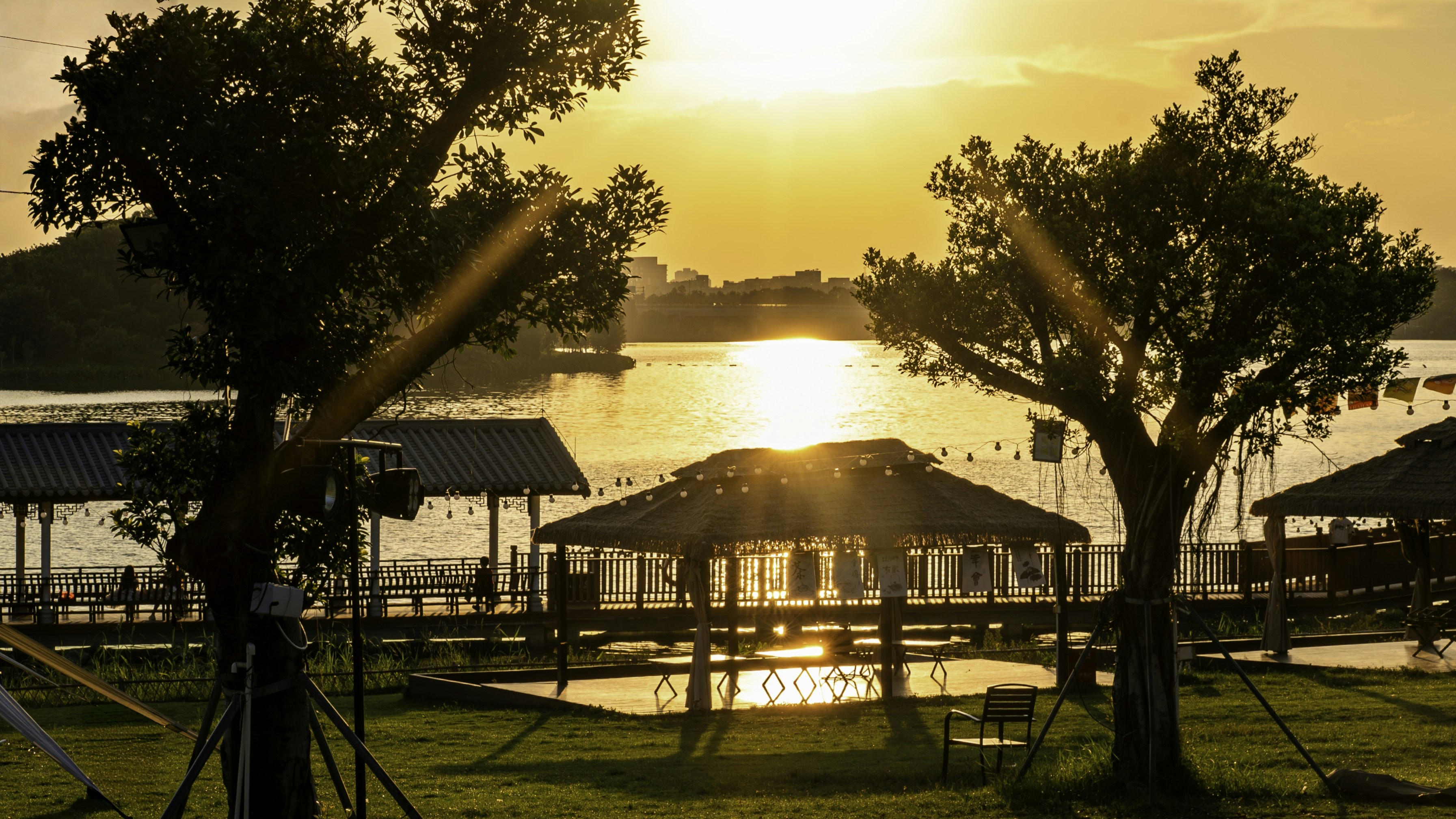 A serene sunset casts a warm, golden glow over a tranquil lake. Silhouetted trees frame the view of the water and lakeside gazebos, with a brilliant sun flare adding a touch of magic to the peaceful evening scene. | Sunset over a lake with trees and gazebos.