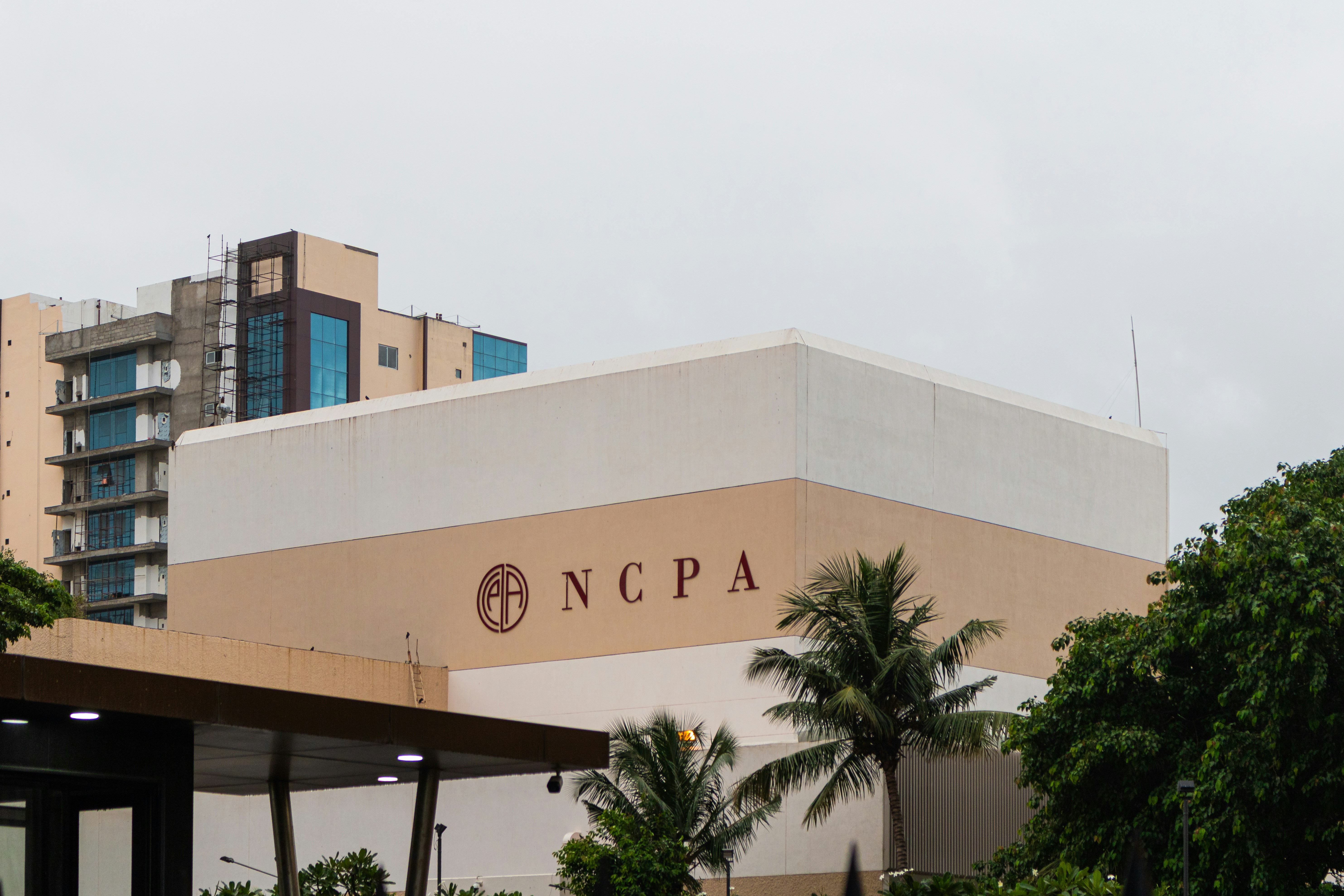 The NCPA building stands prominently amidst urban surroundings, showcasing its distinctive architecture and branding. Lush greenery contrasts with the modern structures nearby.