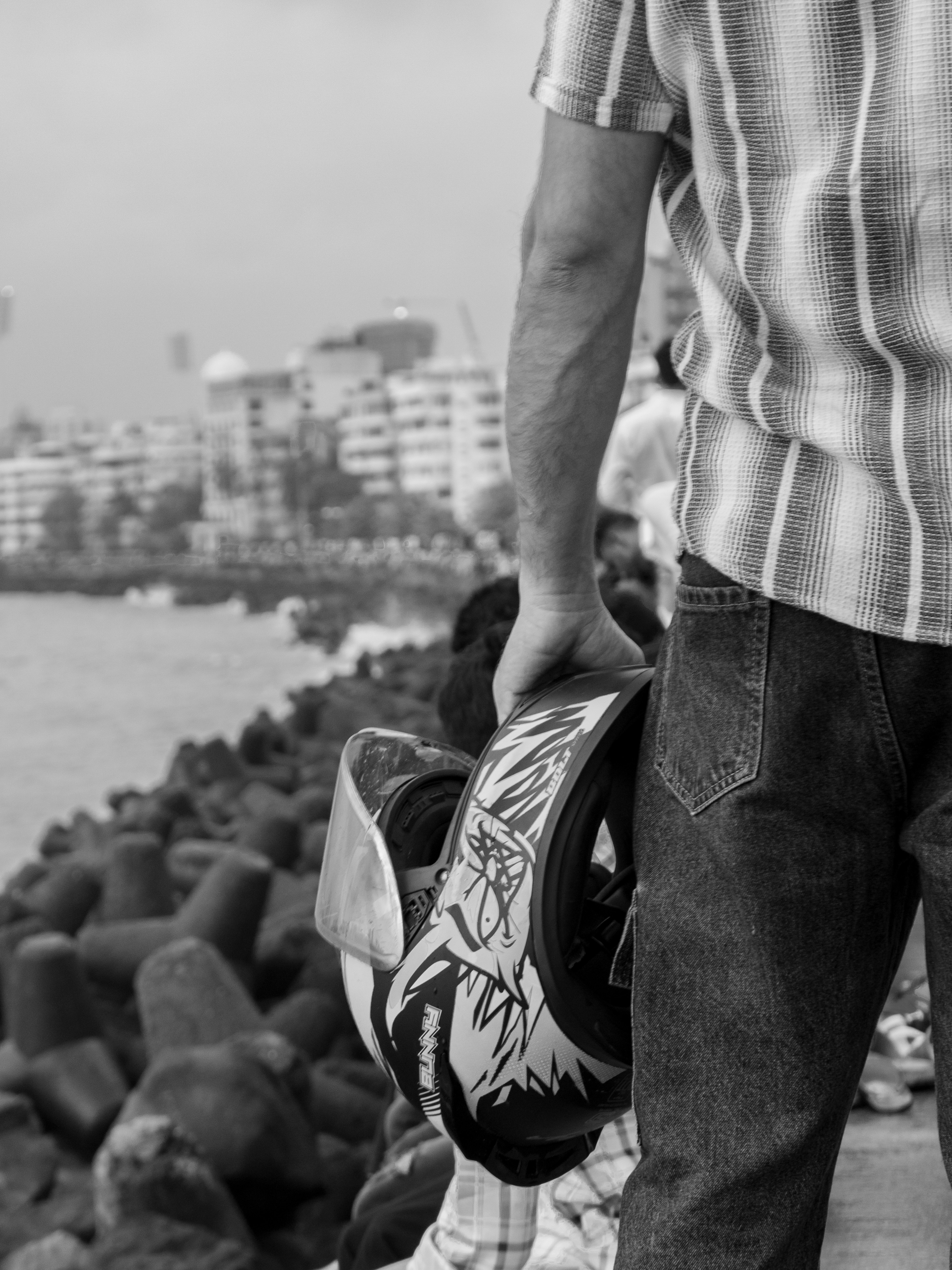 A person holding a motorcycle helmet stands along a rocky shore, gazing at the distant cityscape. The monochrome tones emphasize the contrast between the individual's solitude and the bustling background.