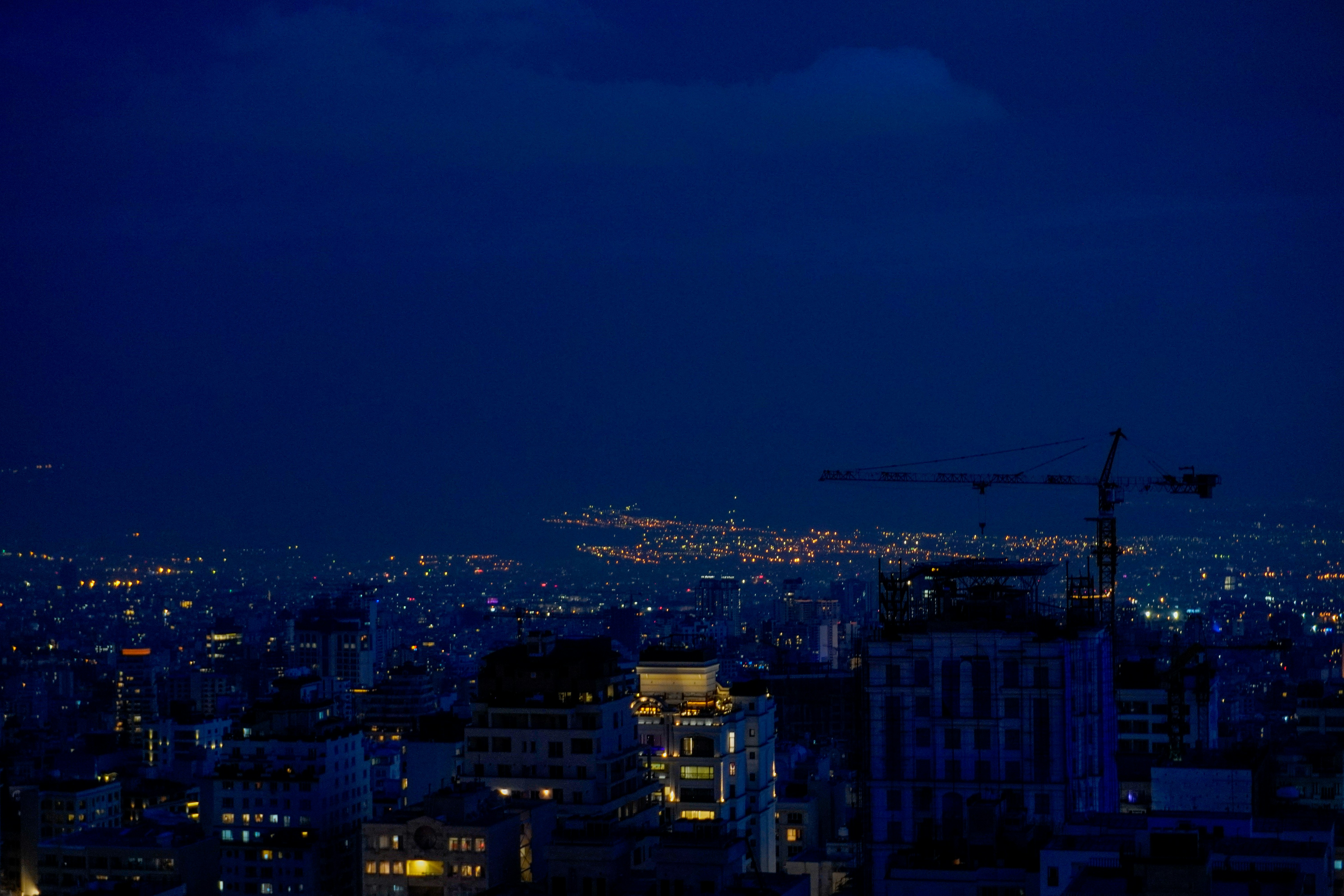 A panoramic view of a cityscape at twilight, showcasing the glow of city lights against a deep blue sky, with construction cranes silhouetted in the foreground.