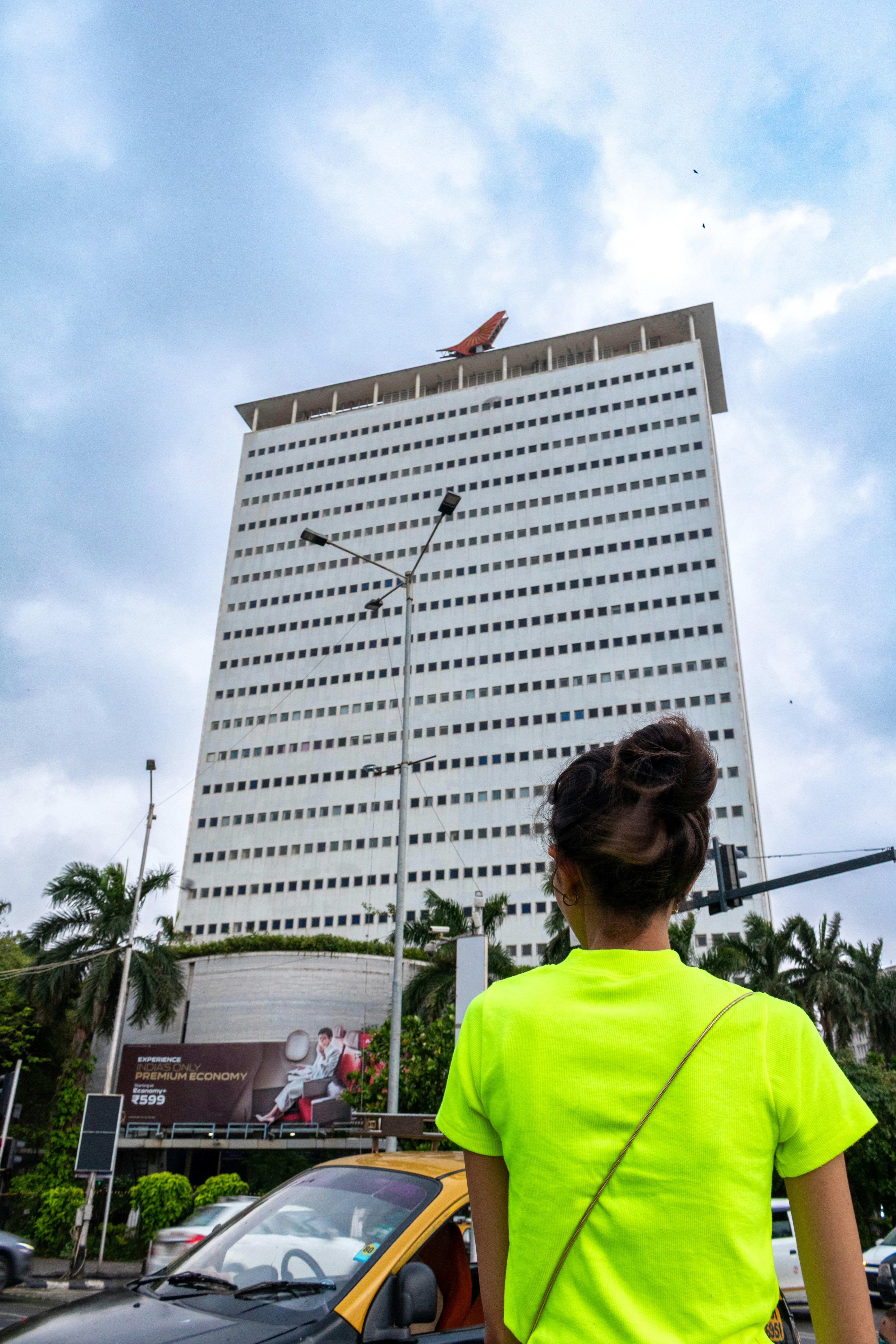A girl wearing a fluorescent green t-shirt stands with her back to the camera, gazing up at the towering Air India Building in Nariman Point, Mumbai. The vivid pop of her shirt contrasts against the neutral tones of the concrete skyscraper, creating a strong visual focal point. | Woman gazes at building with an airplane on top.