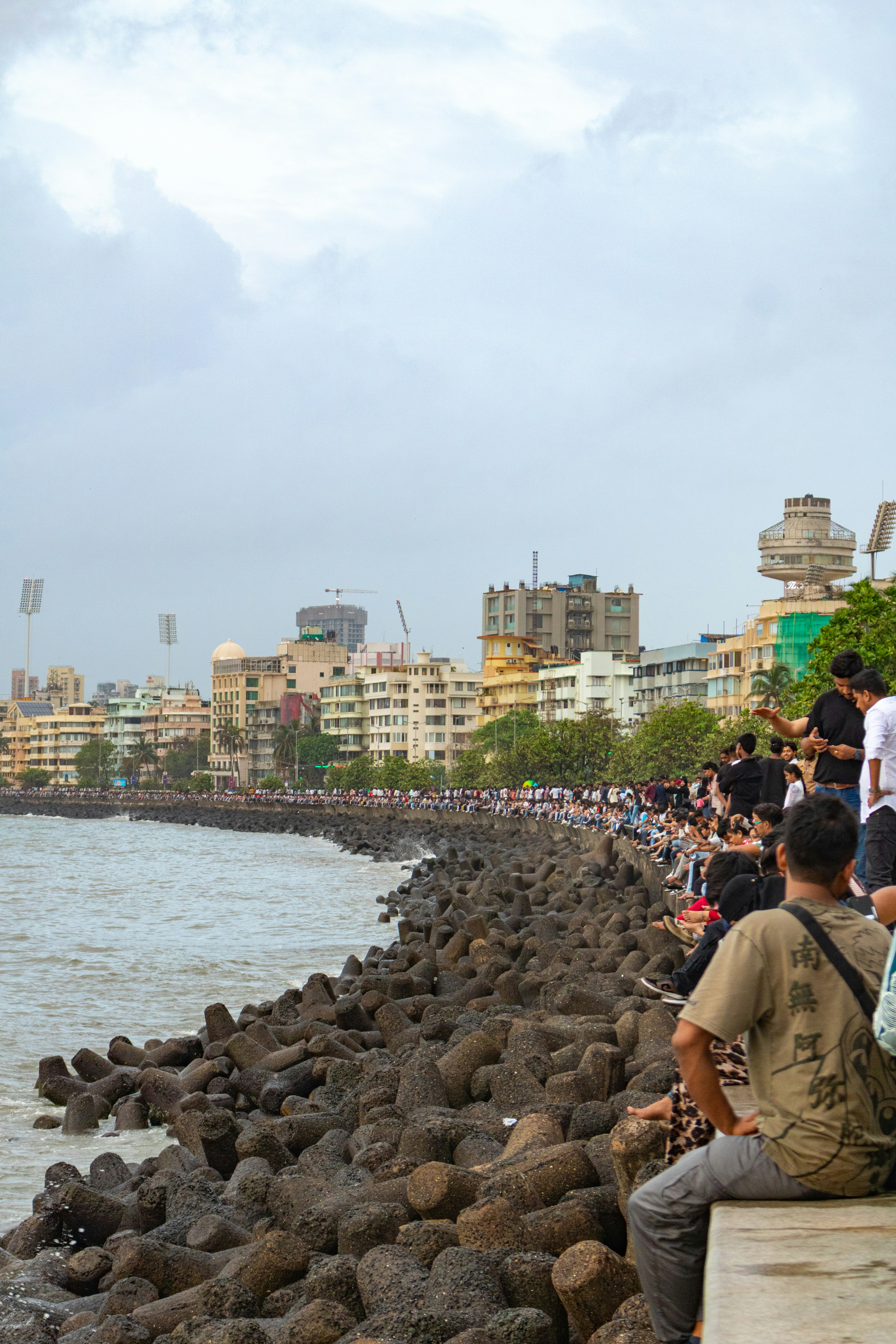 This photo captures the vibrant atmosphere along Marine Drive in South Mumbai, with crowds gathered on the promenade beside the iconic tetrapods. The Arabian Sea rolls in gently, while a dense line of mid-century and modern buildings forms the city’s coastal backdrop. Taken during the monsoon season, the image showcases the layered character of Mumbai — energetic, resilient, and deeply tied to its seafront. With people seated along the edge enjoying the view, this photo highlights the everyday rhythm of life at one of the city’s most loved public spaces.