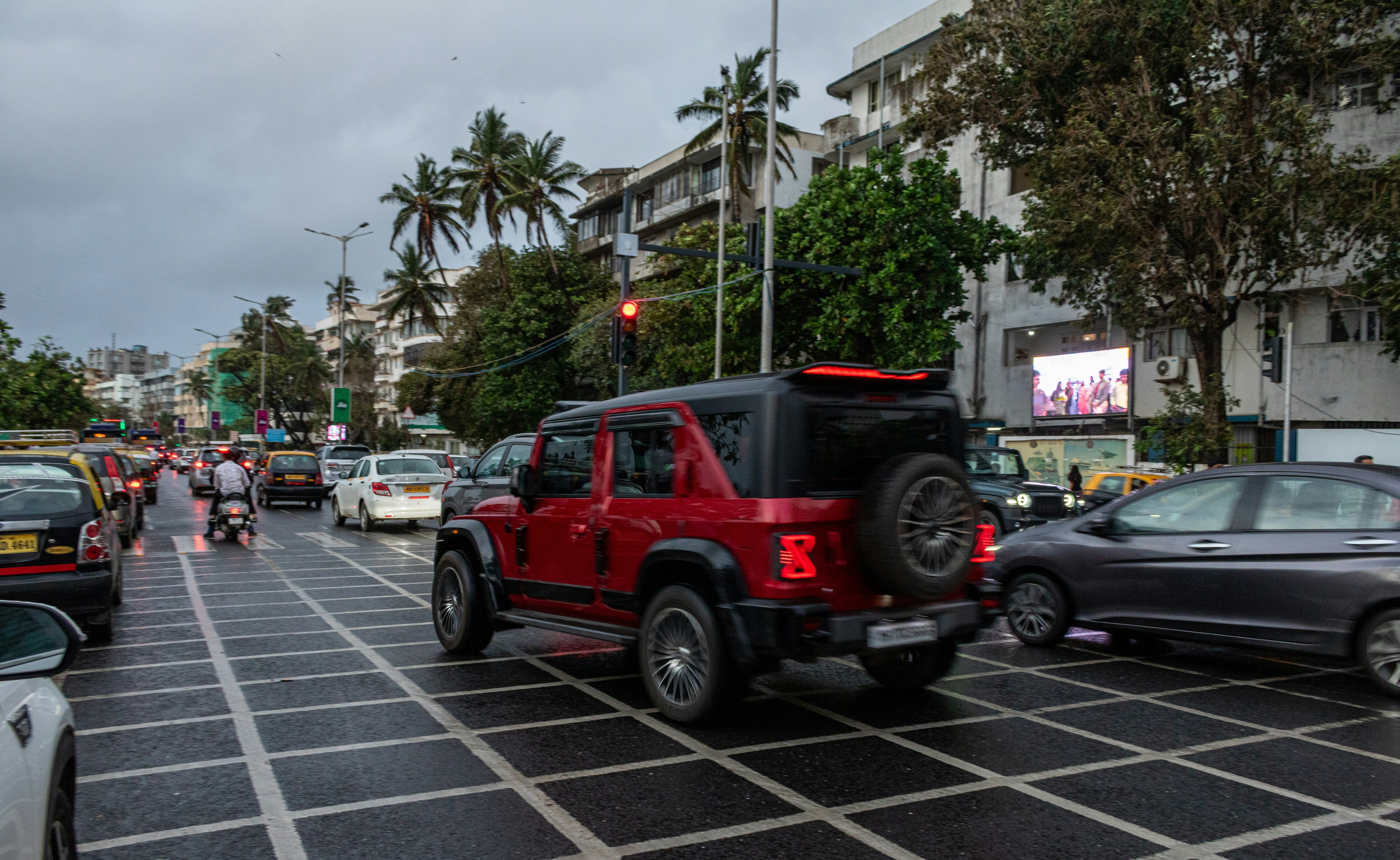 A red Mahindra Thar makes a turn onto a main road, its bold color and rugged build standing out against the urban backdrop. Captured in motion, the SUV reflects a blend of off-road capability and city presence, popular across India for its versatility and design. The image highlights the Thar’s distinctive profile, wide stance, and street-readiness as it joins the flow of traffic. T | A red suv is driving on a wet road.