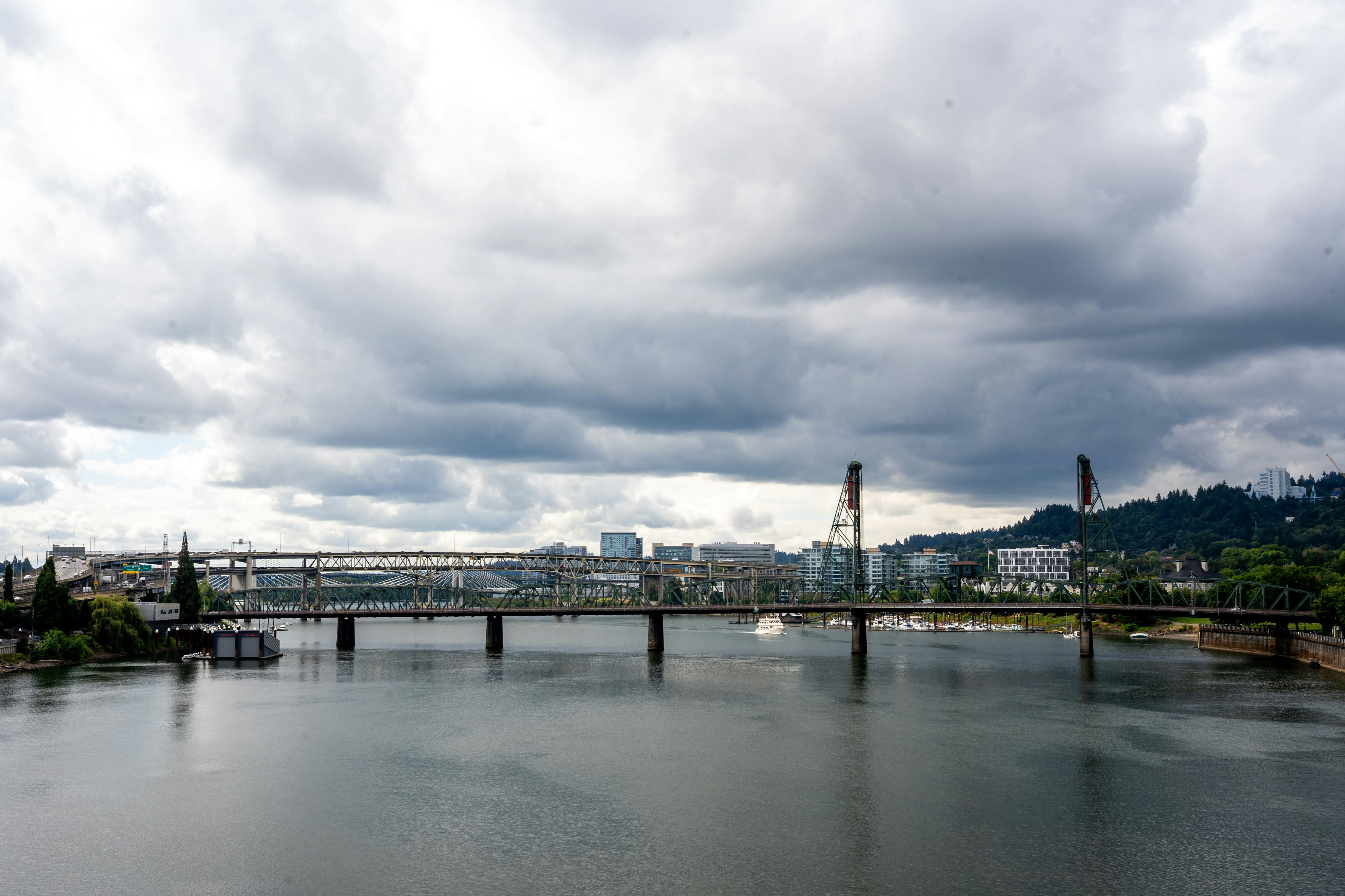 North-facing view from the east side of the Hawthorne Bridge in Portland, Oregon. The photo shows the Willamette River with the Burnside, Steel, and Broadway Bridges in the distance. Mountains are visible on the horizon, and Portland’s waterfront areas line both sides of the river. | A bridge spans over a calm river.