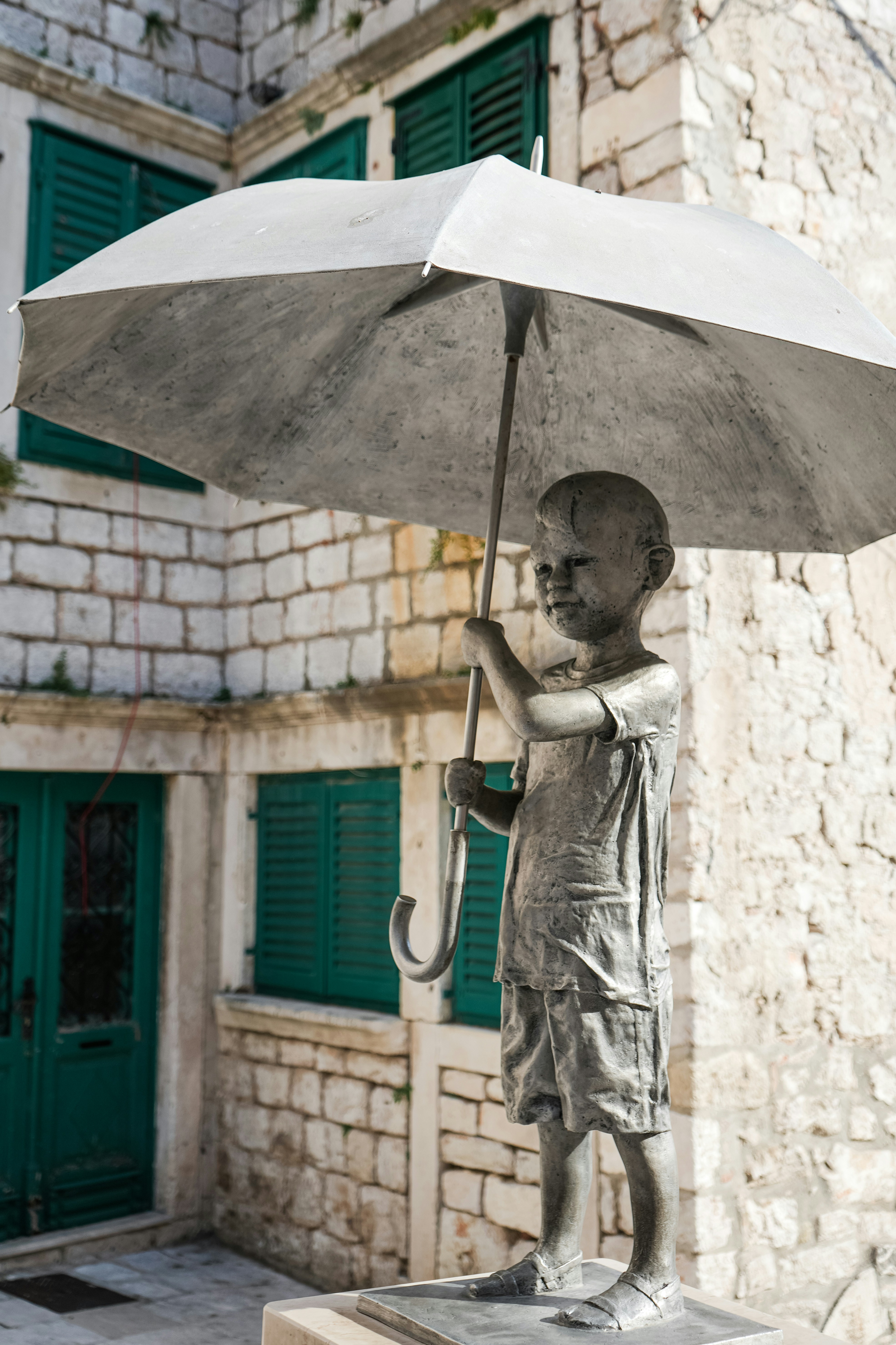 Bronze statue of a child holding an umbrella, set against a backdrop of stone walls and green shutters.