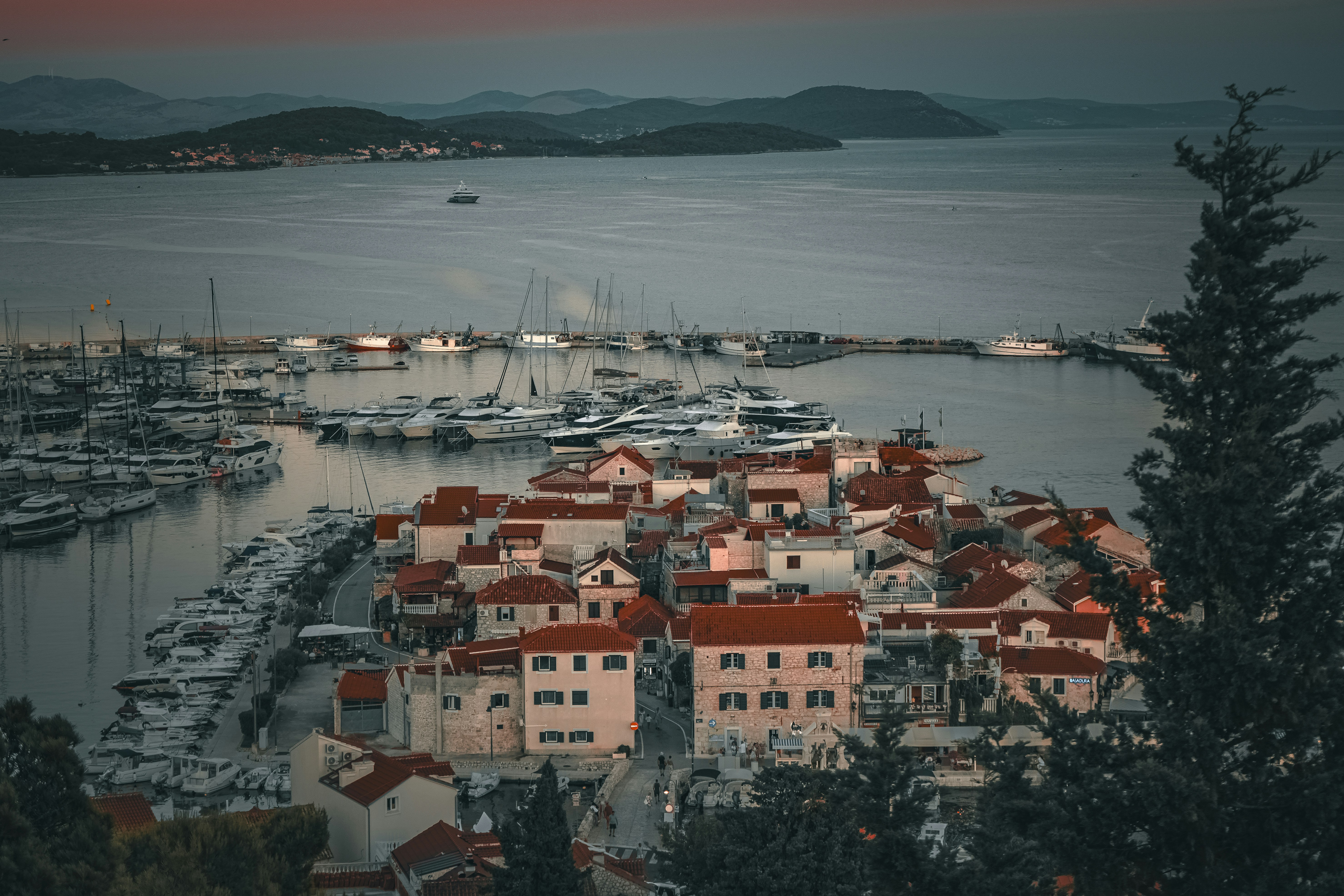 A picturesque coastal village nestled by a marina, showcasing charming red-roofed houses and moored yachts under a twilight sky.