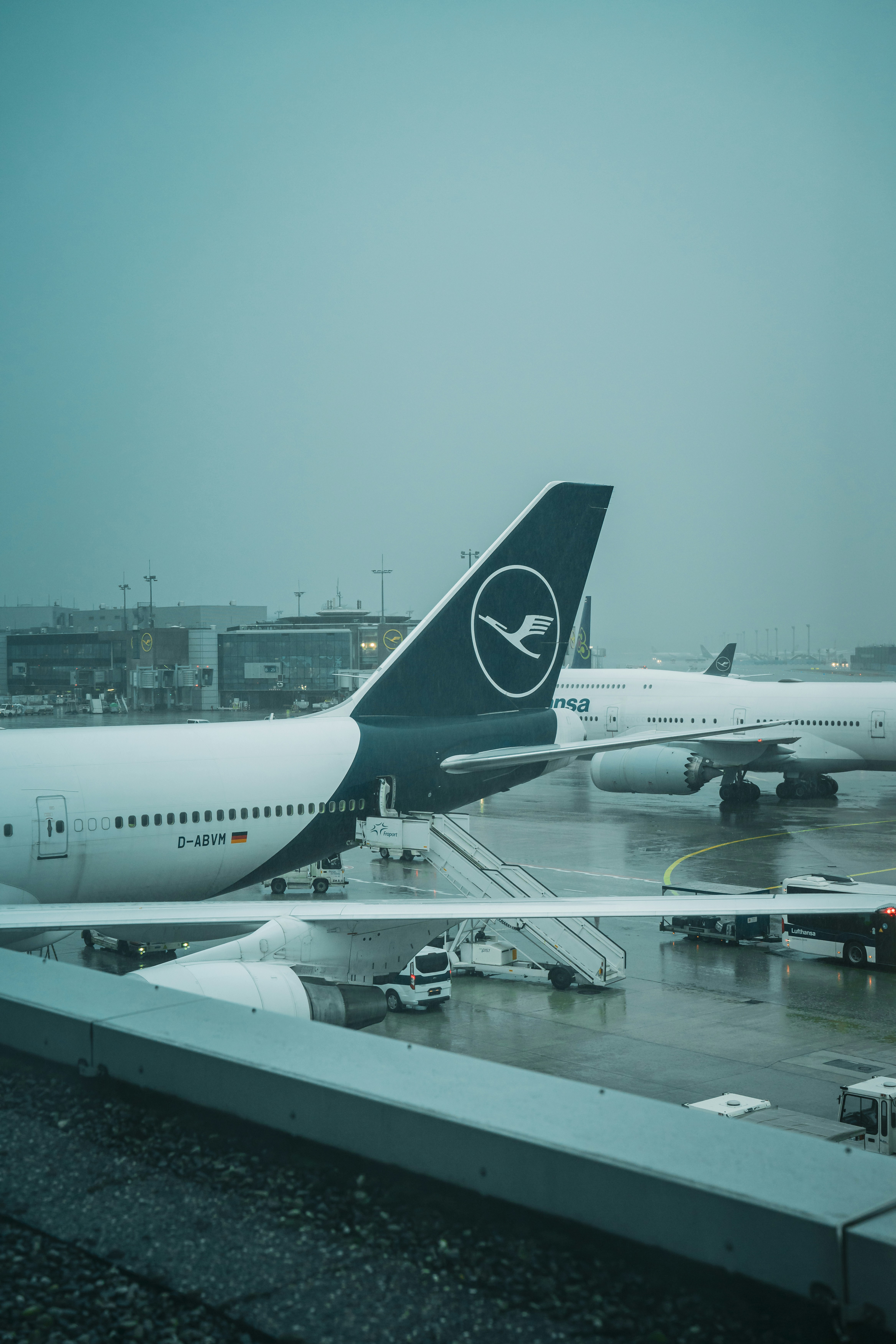 Lufthansa aircraft parked at a rain-soaked airport, with a focus on the distinct tail design. The scene captures the essence of travel amidst gloomy weather.