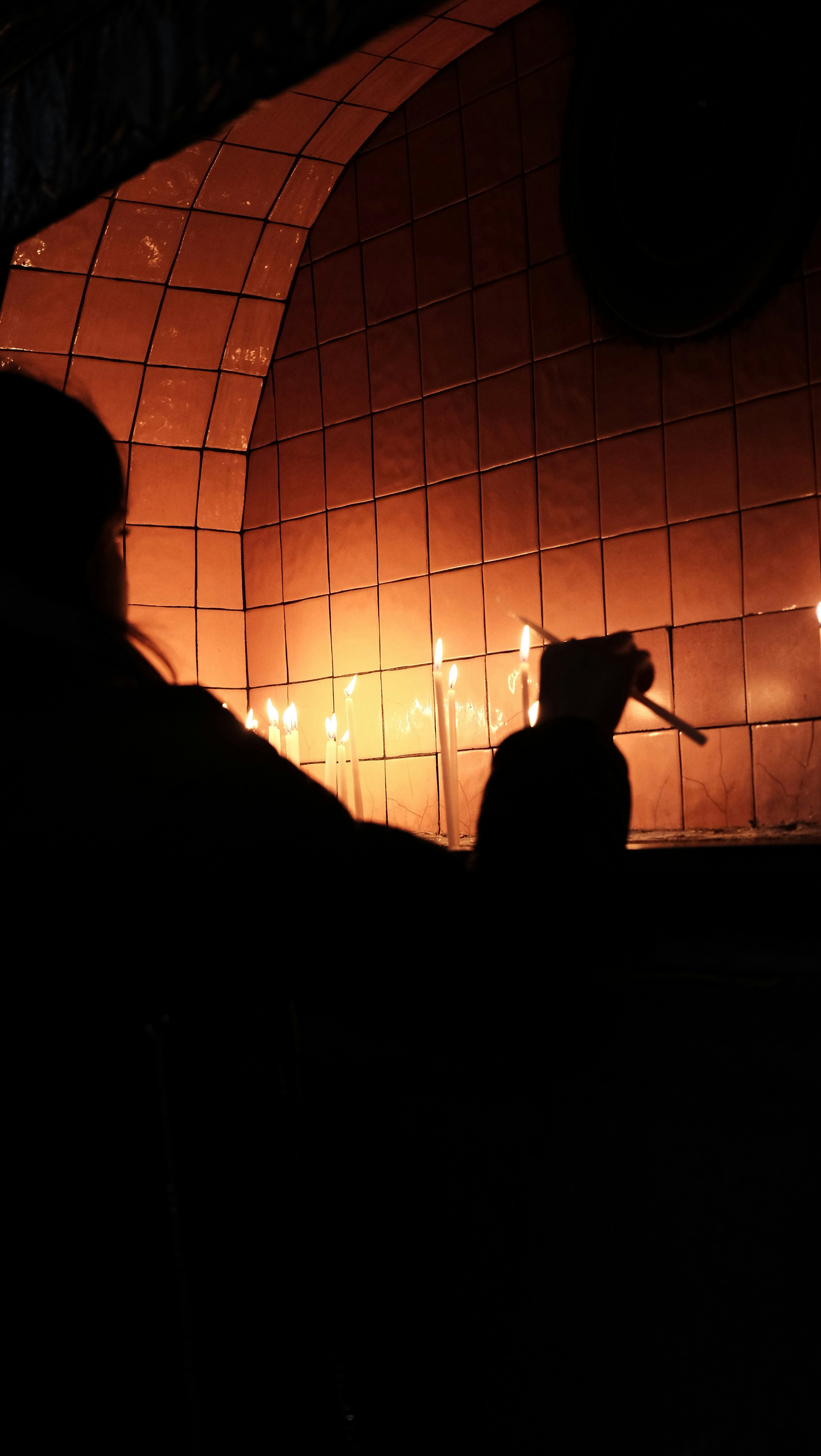 A quiet moment of reverence as a figure lights a candle among many in a softly glowing prayer alcove. The warm reflections on the tiled wall and the gentle flicker of flame create an atmosphere of contemplation, hope, and sacred stillness — a timeless ritual captured in a single frame. | Someone is lighting candles in the dark.