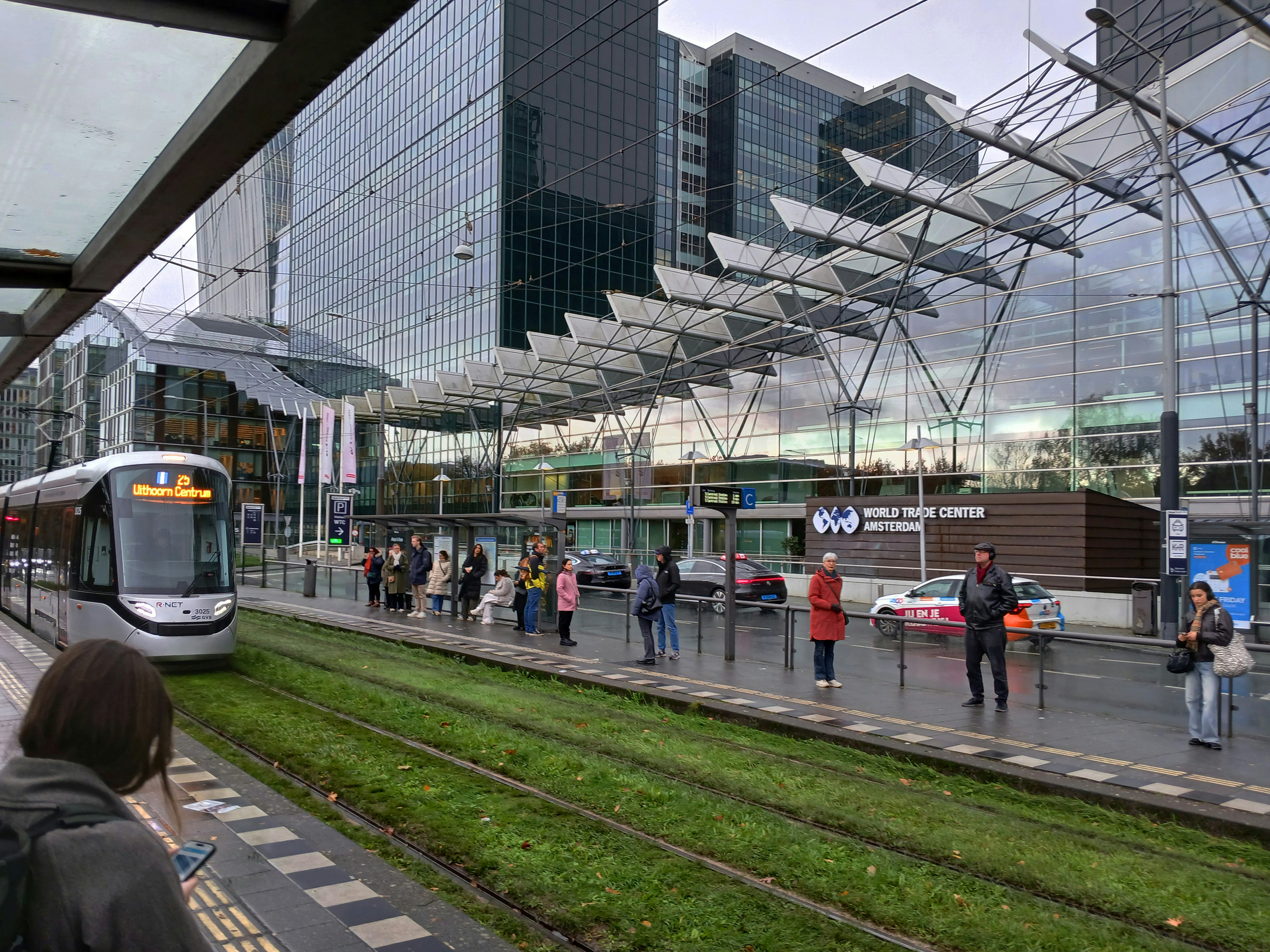 Free download photo of tram track and platform where city people are waiting, with in the background modern architecture of the so-called Zuid-As in city Amsterdam. I like the reflections in all the glass walls and roofs. A grey day in February. Street photography free image in high resolutions by Fons Heijnsbroek, The Netherlands Dutch photo / Gratis download foto van tram, trambaan en perron waar mensen wachten, met op de achtergrond de moderne architectuur van de Zuid-As in Amsterdam. Ik vind de reflecties in de glazen wanden en daken prachtig. Een grijze dag in februari. Straatfotografie. | A modern tram pulls into a city station.