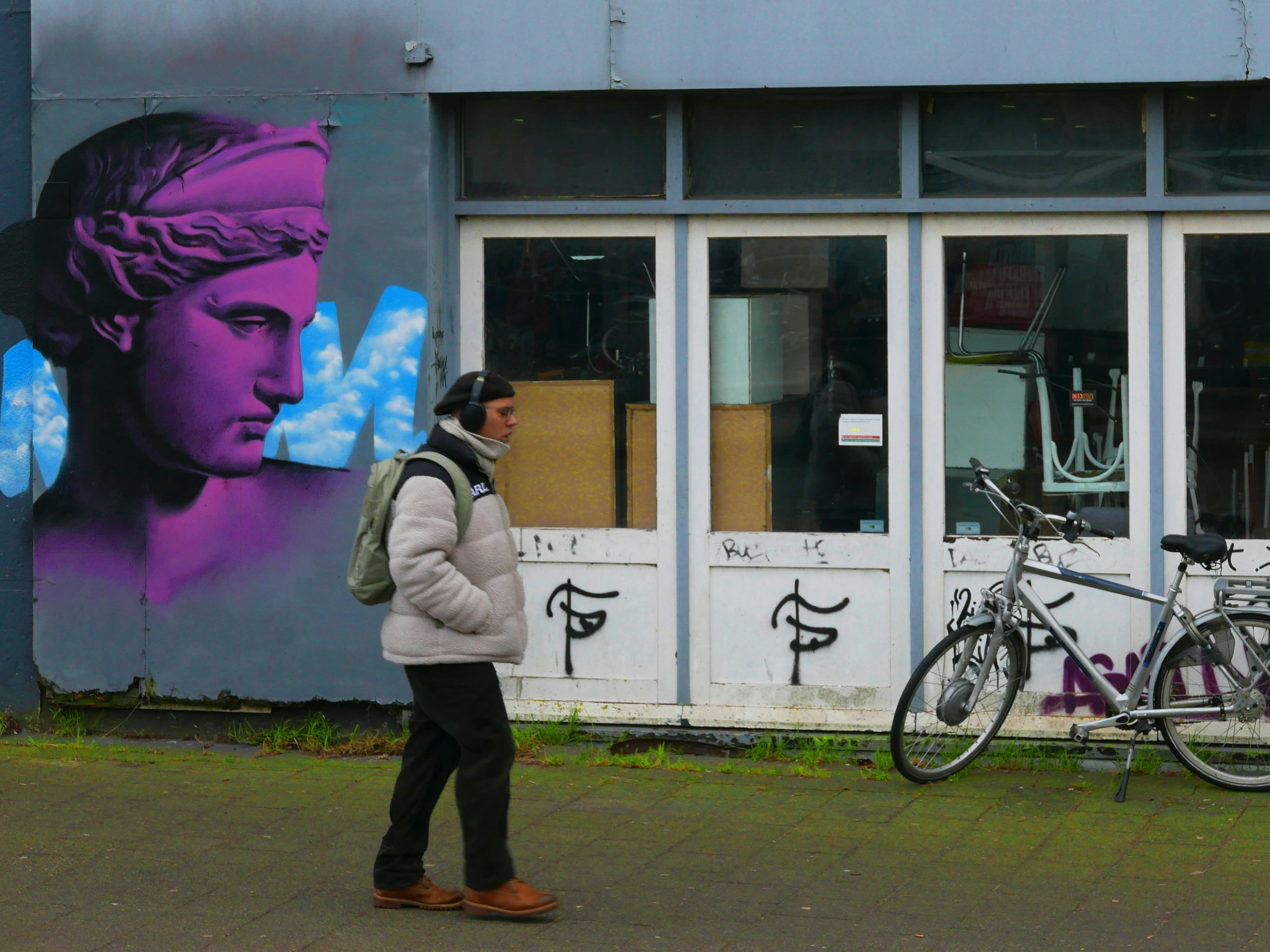 Free download photo of a man walking on the sidewalk with a closed shop in the background and a classical head, painted on the wall. It is in the street Sarphatistraa in city Amsterdam. I like the classcial mural very much, like a city angel! Street photography free image in high resolutions by Fons Heijnsbroek, The Netherlands Dutch photo / Gratis download foto van een man die op de stoep loopt met een gesloten winkel op de achtergrond en een klassiek hoofd, geschilderd op de muur. Het is in de Sarphatistraat in Amsterdam. Ik vind de klassieke muurschildering erg mooi, als een stadsengel! | Street art portrait alongside a walking person.