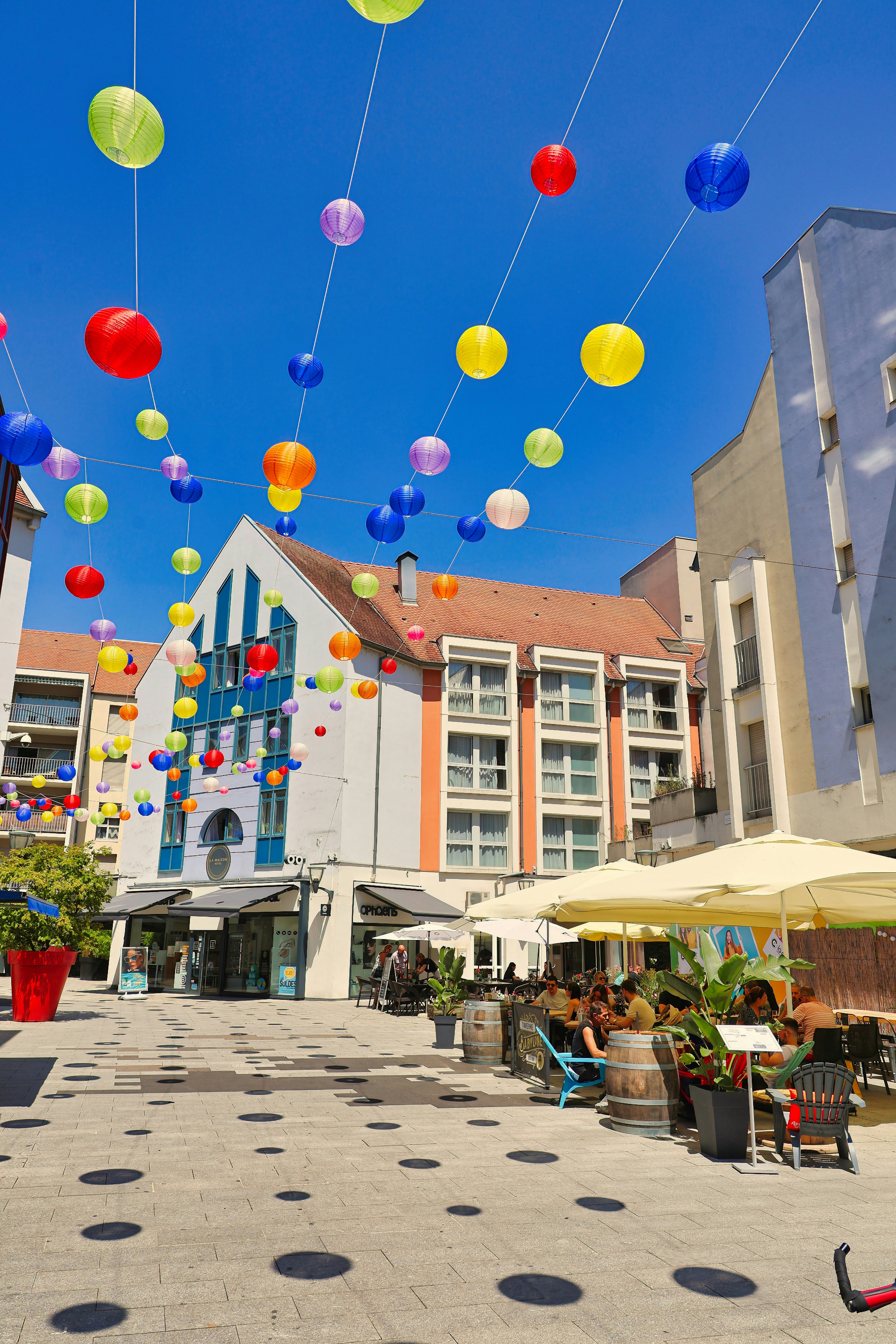 La zone piétonne de Mulhouse s’organise autour d’un axe commerçant animé, qui combine grands magasins et galeries marchandes, et se prolonge par un réseau de petites rues plus calmes, ponctuées de restaurants, de terrasses et de boutiques indépendantes. Elle débouche sur la place de la Réunion, vaste espace monumental bordé de bâtiments historiques. Depuis 2023, la ville a engagé un programme de végétalisation de ces rues piétonnes, visant à renforcer l’agrément et le confort d’usage en période estivale | Colorful lanterns decorate a charming european plaza.