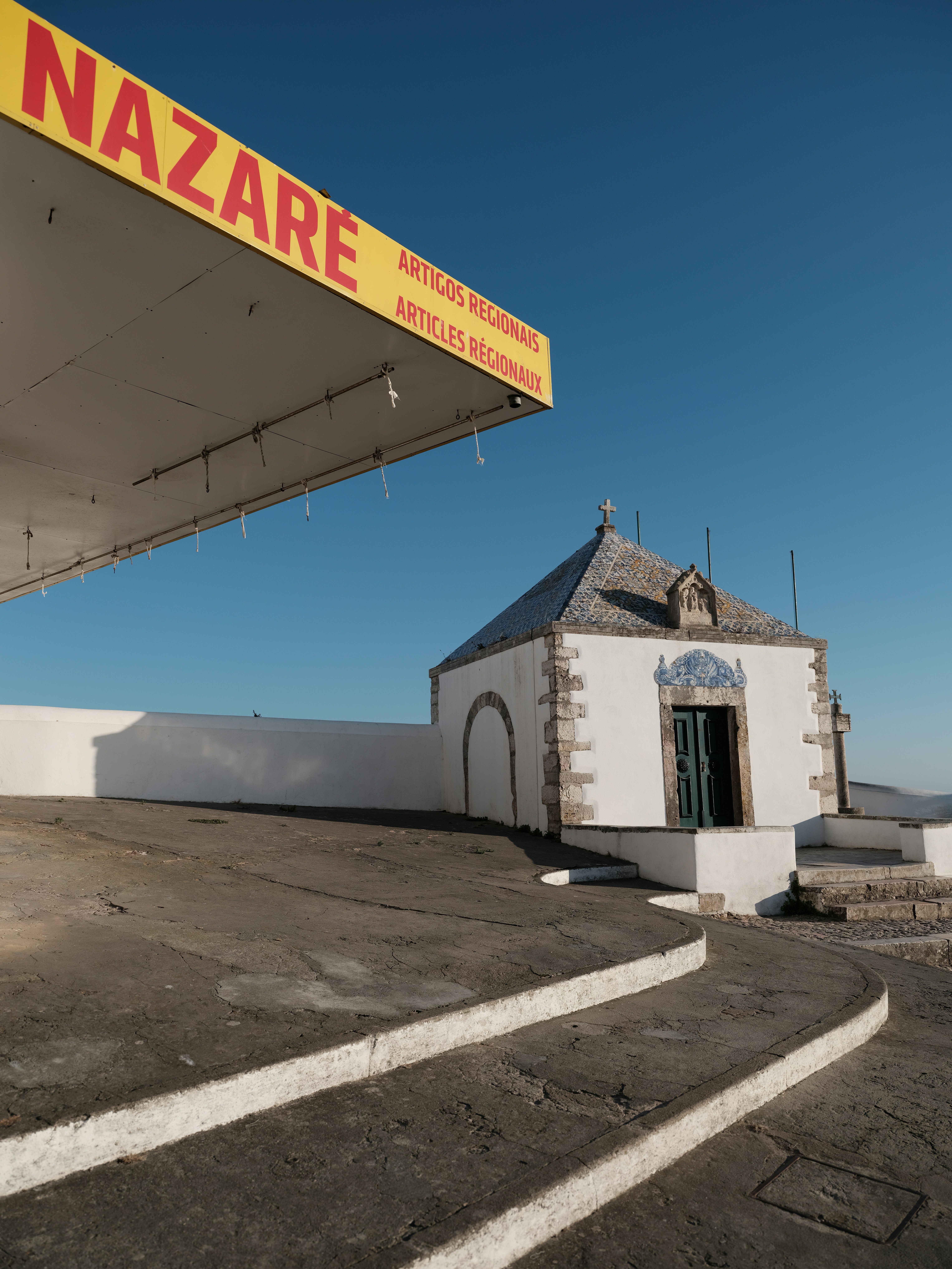 There’s something quietly comic in the way the yellow sign shouts above a landscape that clearly doesn’t care about advertising. I’ve always believed cinema begins in places like this: in silence, in light, in the way architecture and emptiness talk to each other when no one’s around. | A small chapel sits under a nazaré sign.