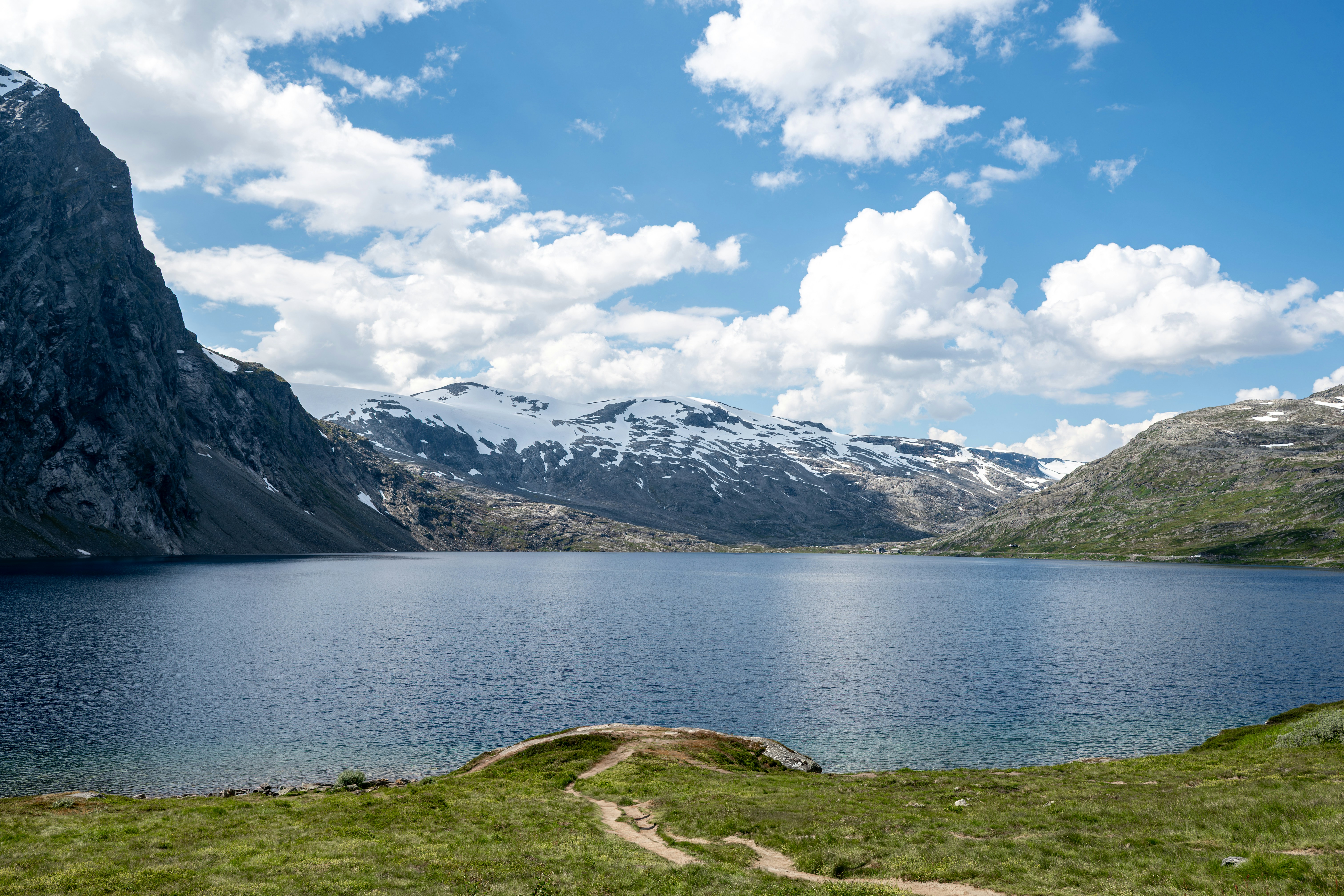 Hiking along the shores of a pristine alpine lake surrounded by snow-capped peaks in Norway's highlands. | A lake reflects mountains under a sunny sky.