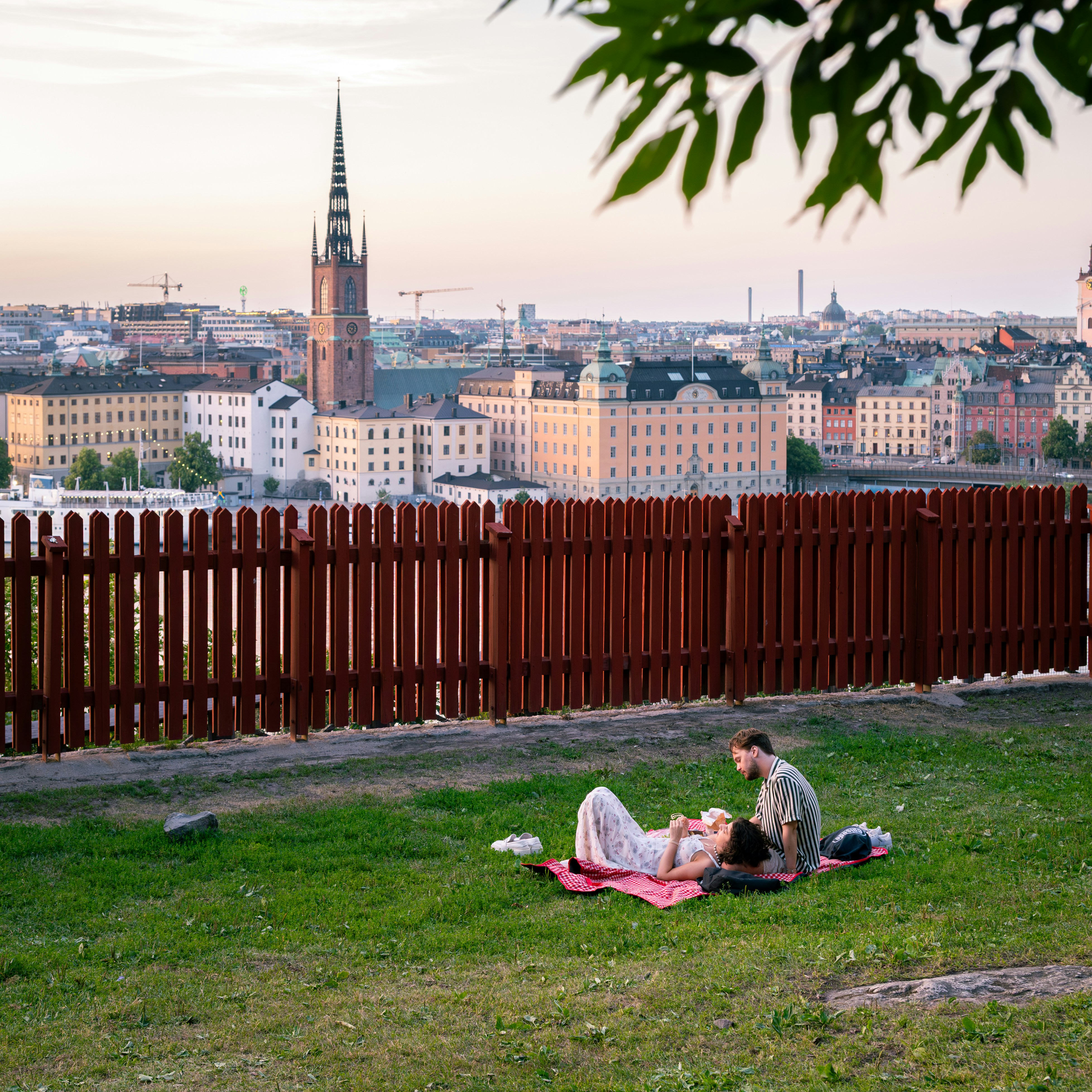Couple relaxing on a blanket in a grassy area, overlooking a picturesque cityscape at dusk with historic architecture and a tranquil river.