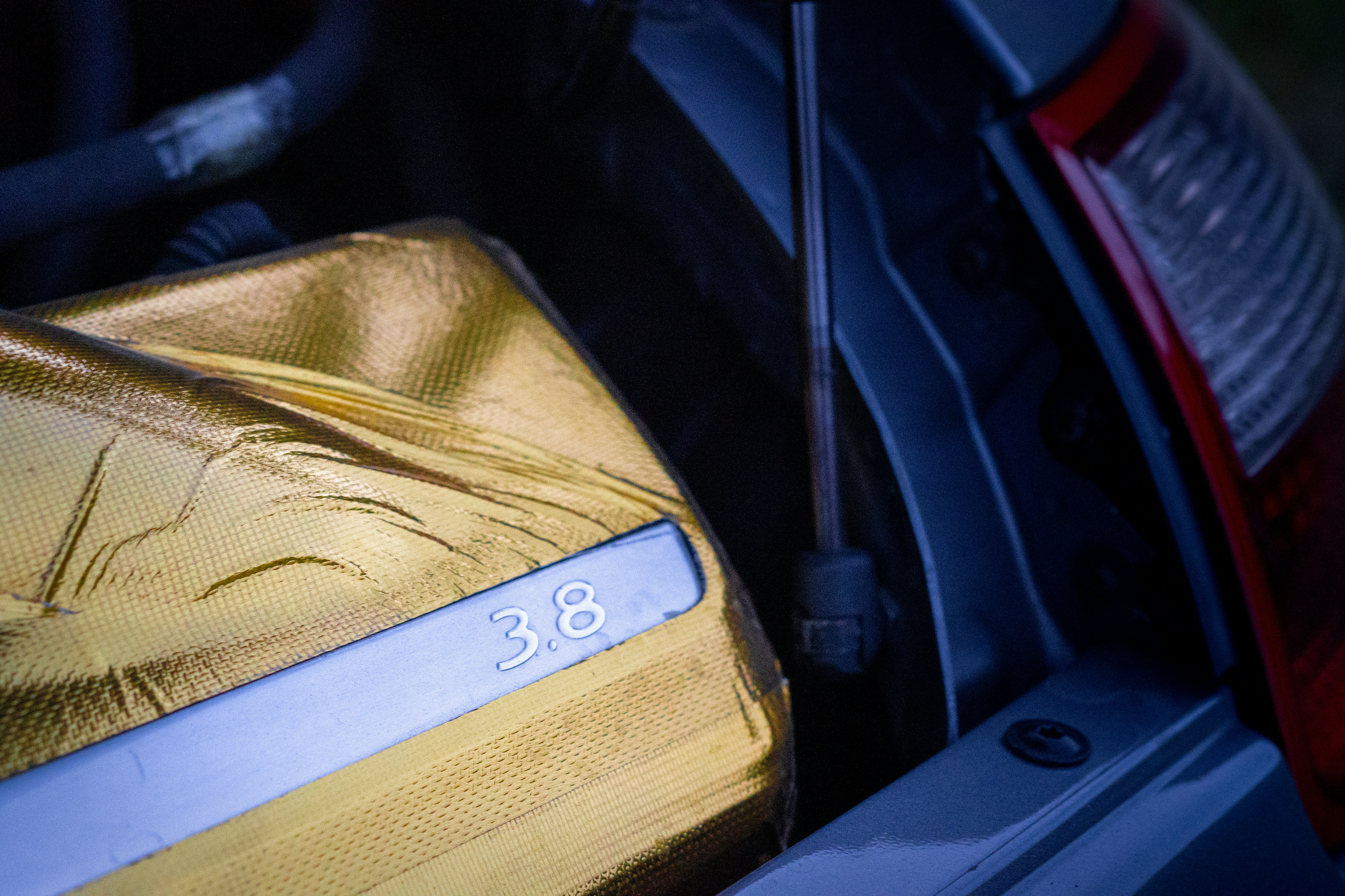 Close-up of a gold-wrapped engine component, revealing the number 3.8, with parts of a car's interior visible in the background.