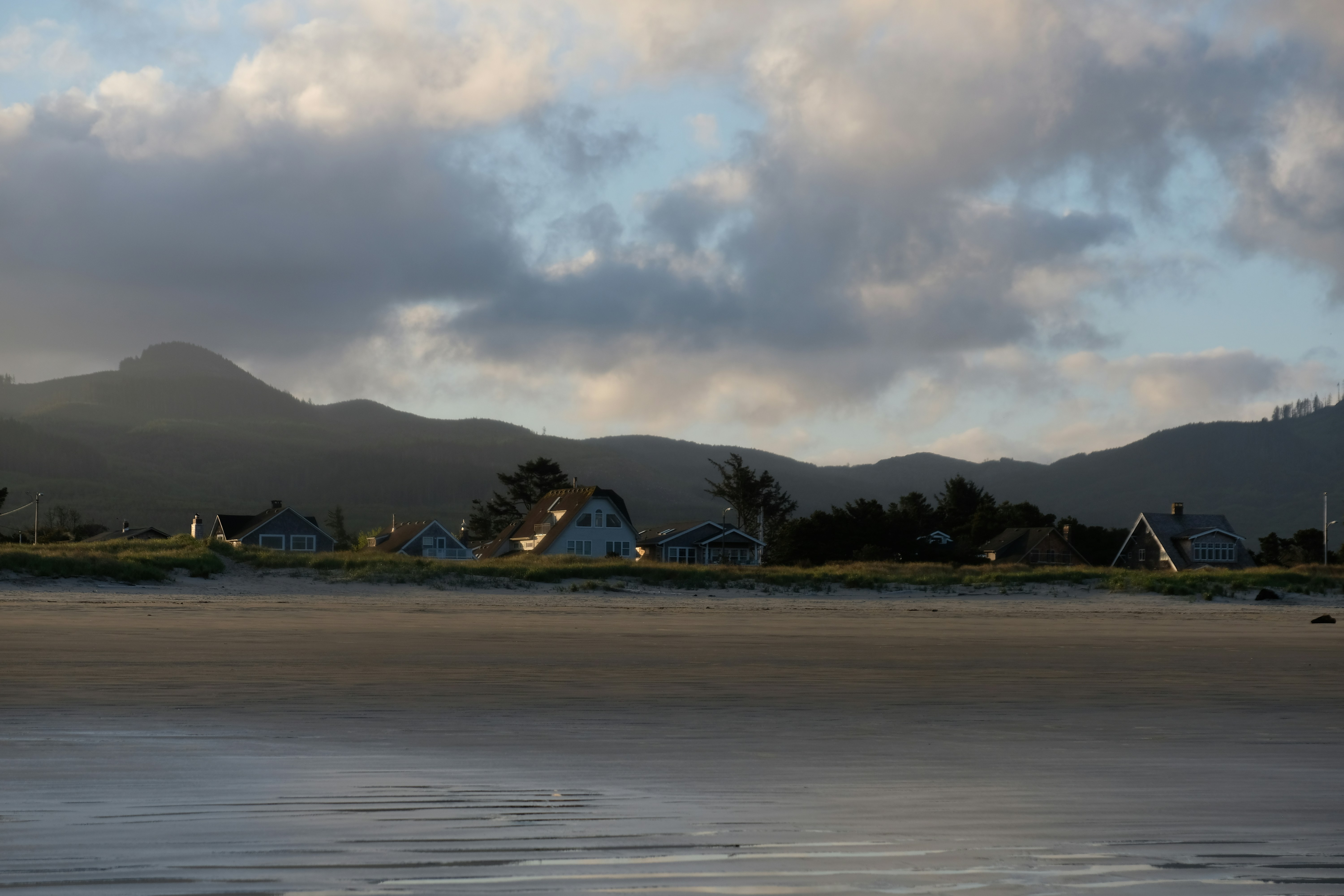 Charming beach houses nestled against a backdrop of rolling hills and cloudy skies. The tranquil shoreline reflects the soft light of the evening.